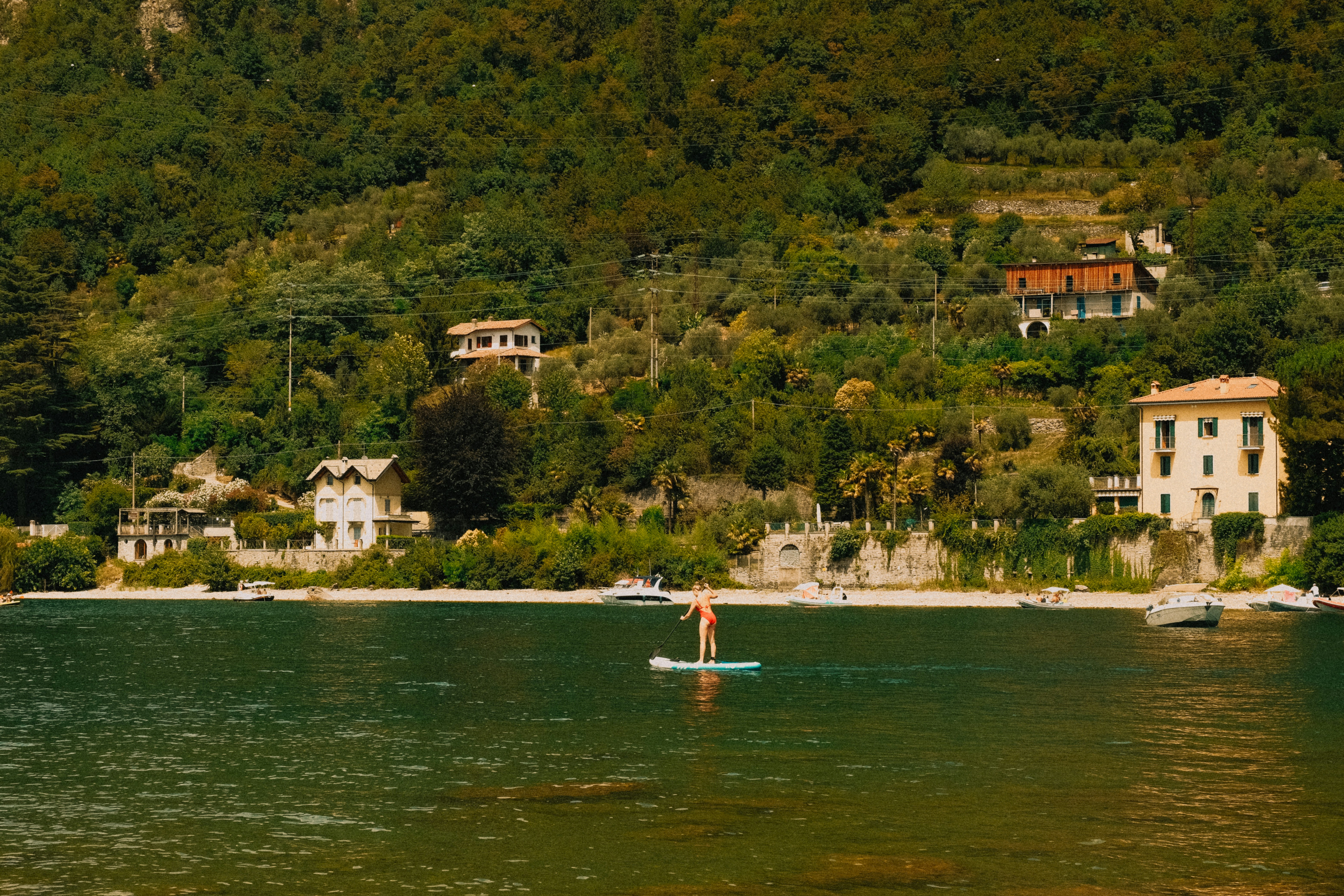 A paddleboarder glides effortlessly across a calm lake, surrounded by lush greenery and charming hillside homes. The tranquil scene captures a moment of leisure in nature.