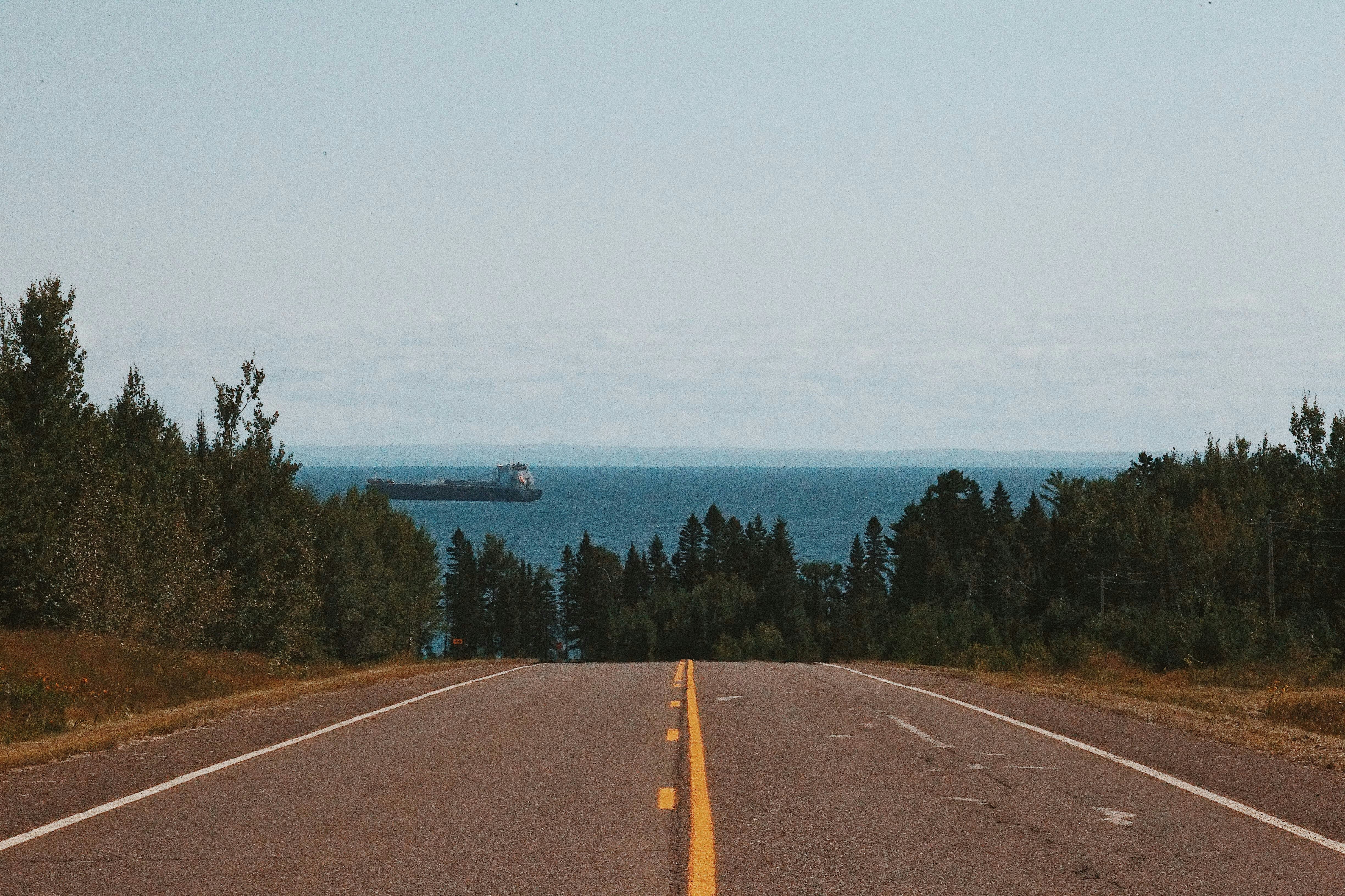 A winding road leads towards the horizon, flanked by lush trees, with a distant cargo ship navigating the serene waters beyond.
