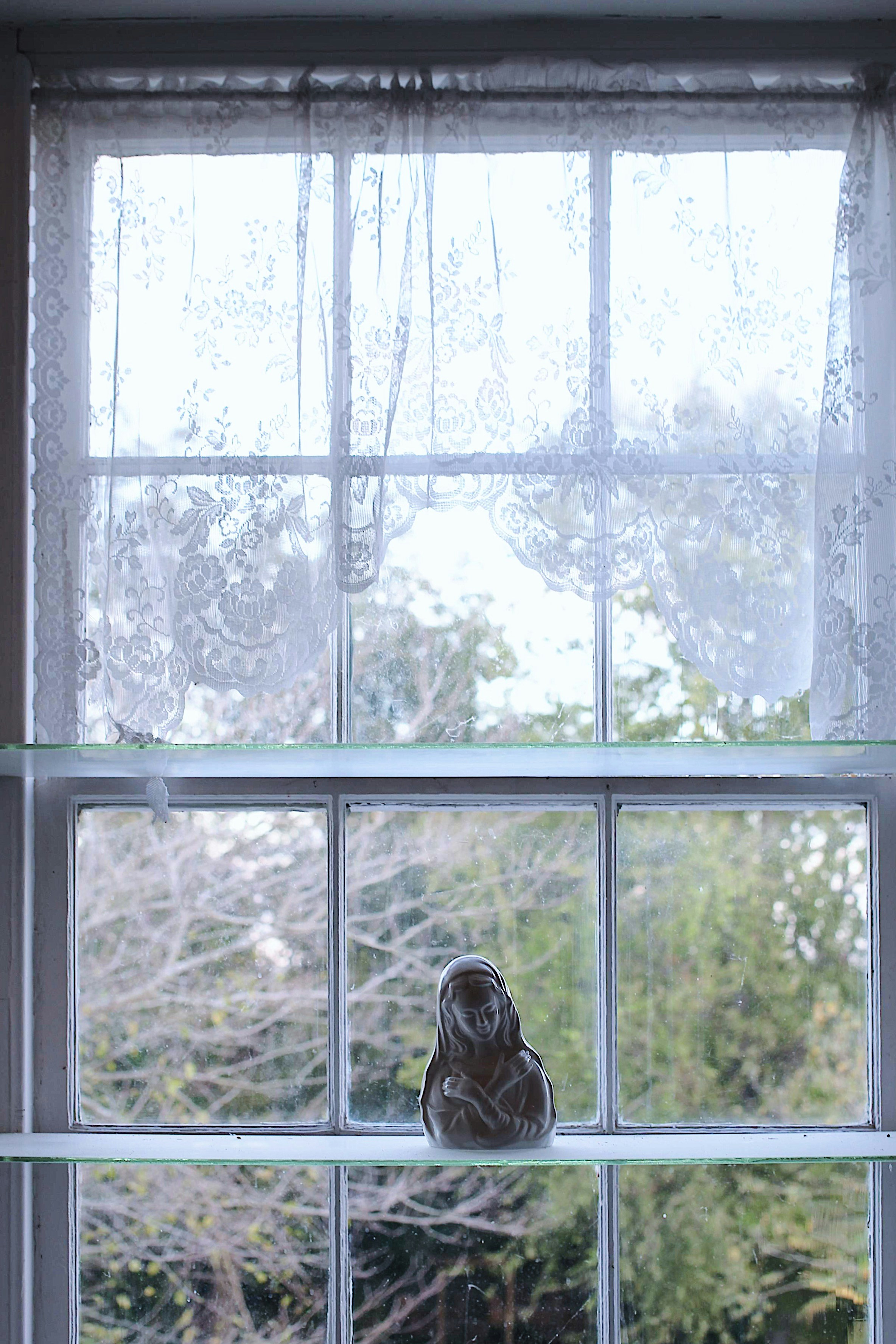 A delicate lace curtain filters light over a serene window scene, featuring a small statue on the sill with greenery visible outside.