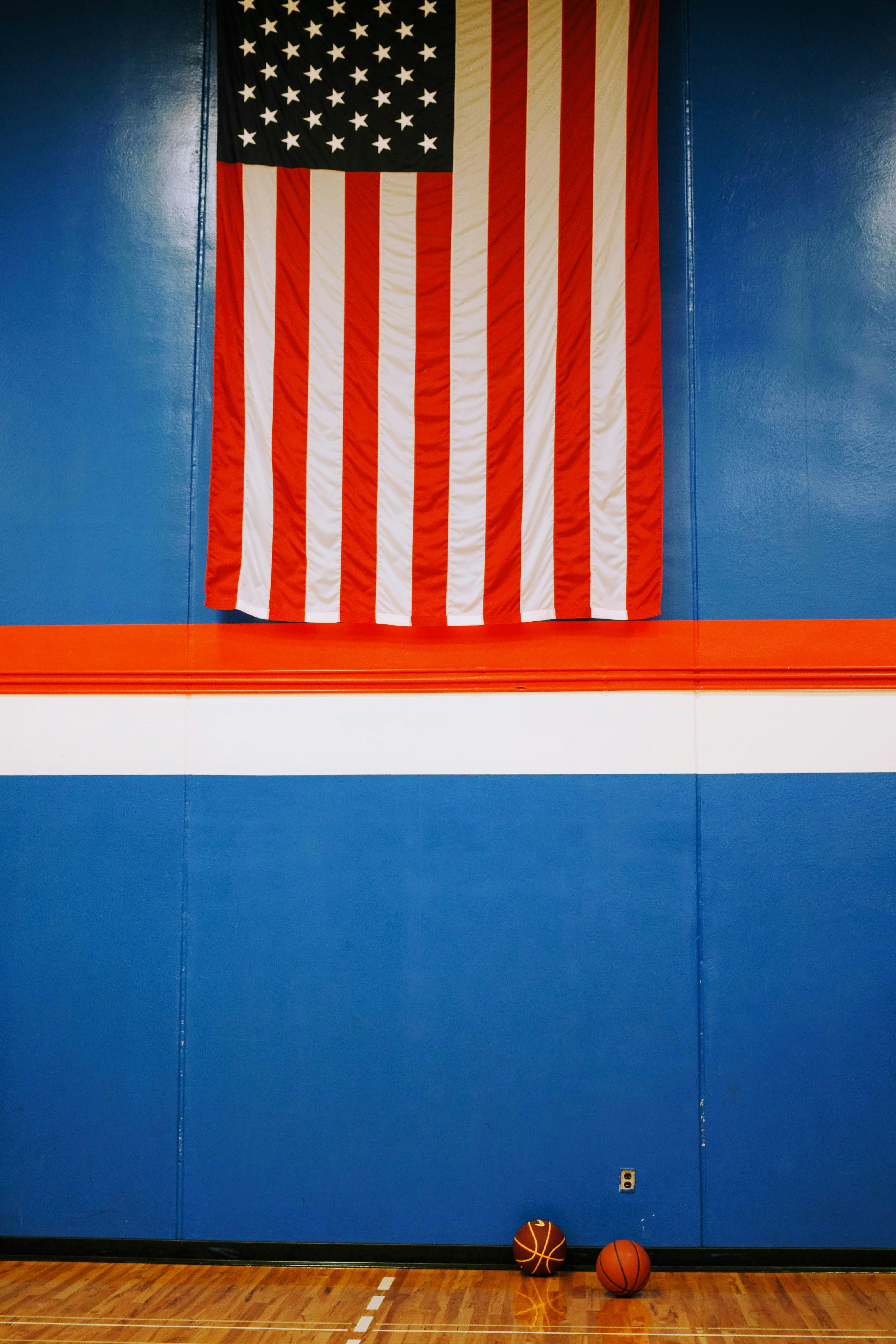 American flag prominently displayed on a vibrant blue wall above two basketballs resting on a gym floor.