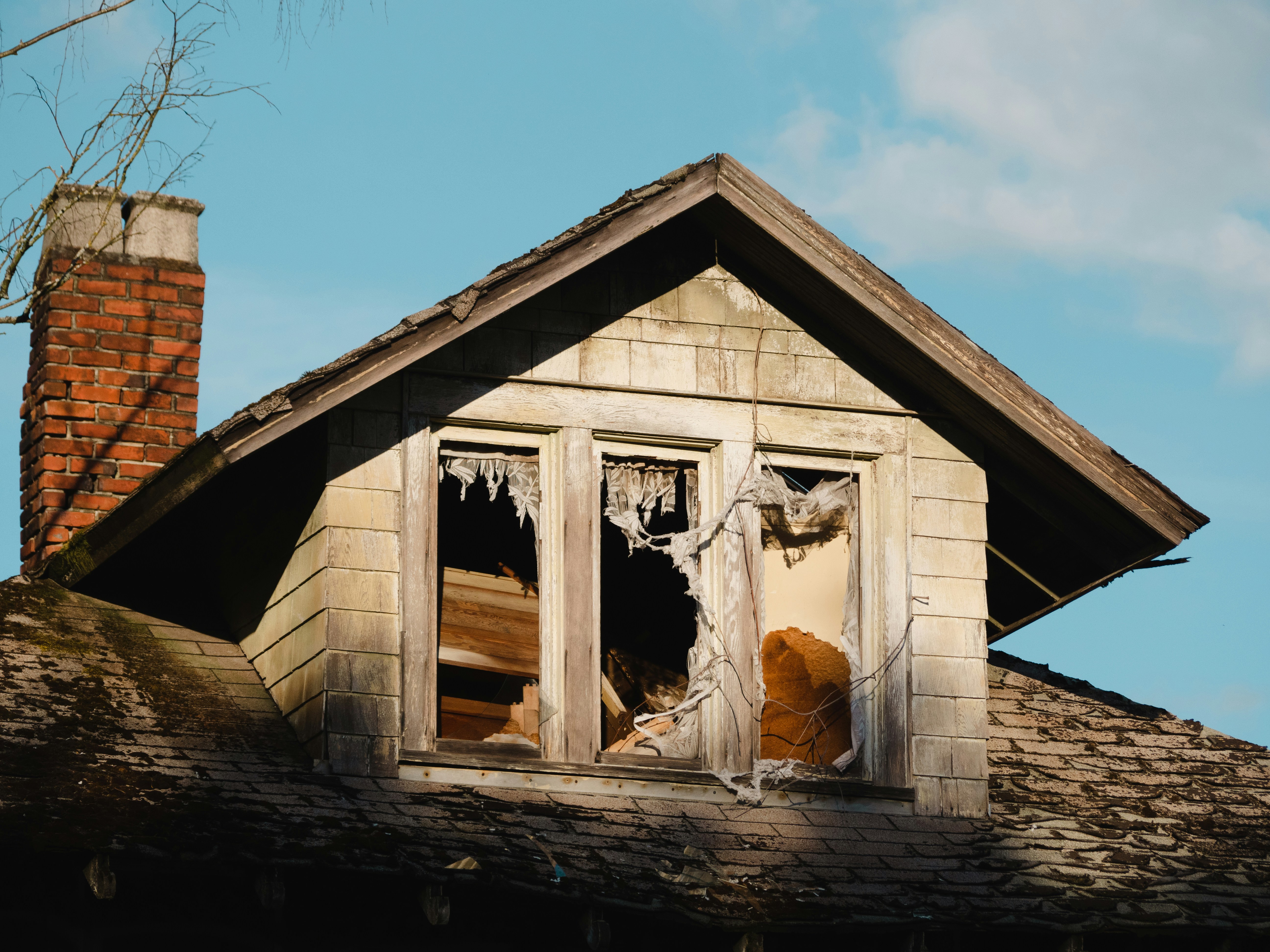 Dilapidated house with broken windows and peeling paint, revealing the remnants of its past. The sunlight casts shadows on the weathered roof.