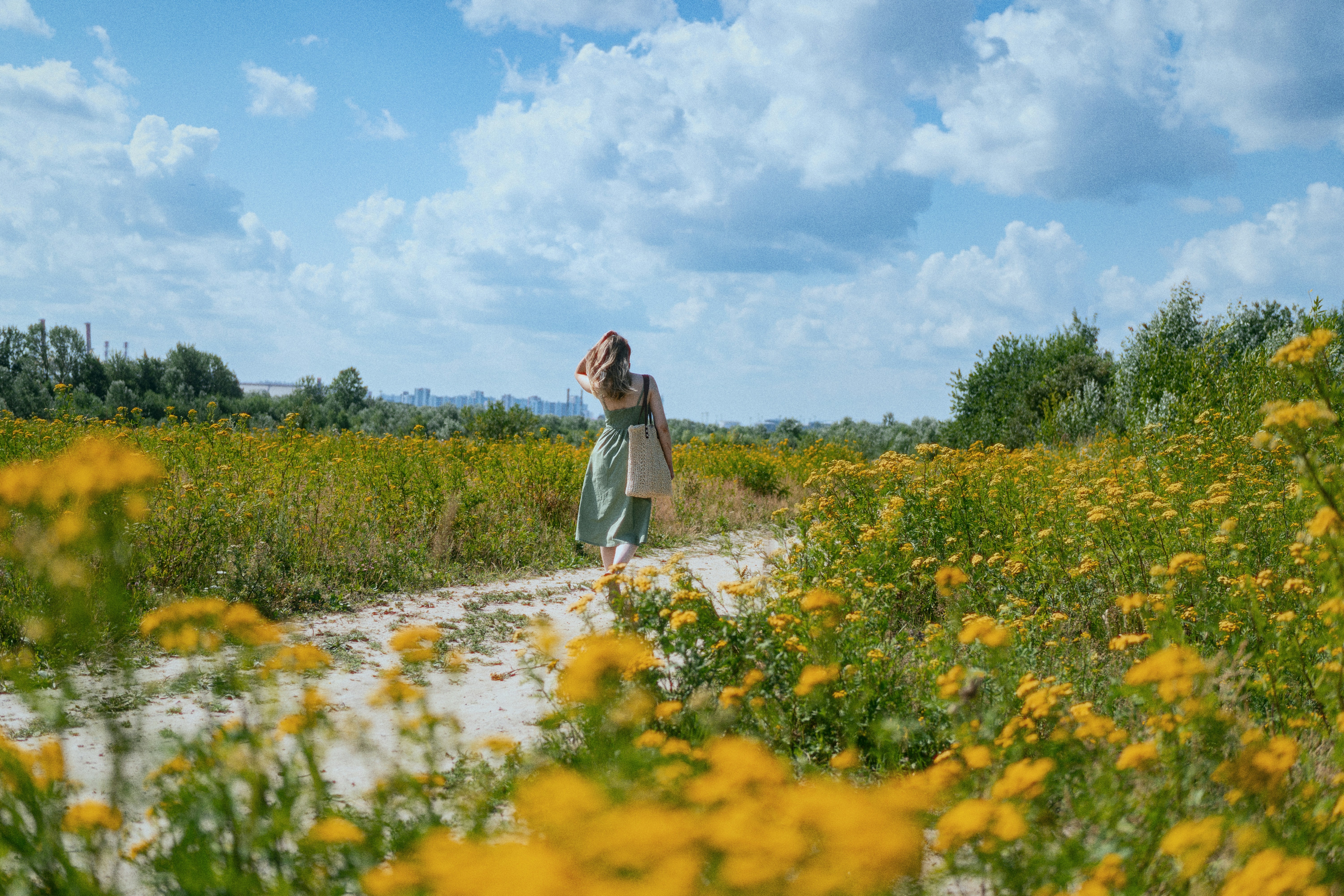A woman strolls along a dirt path surrounded by vibrant yellow flowers under a bright blue sky with fluffy clouds.