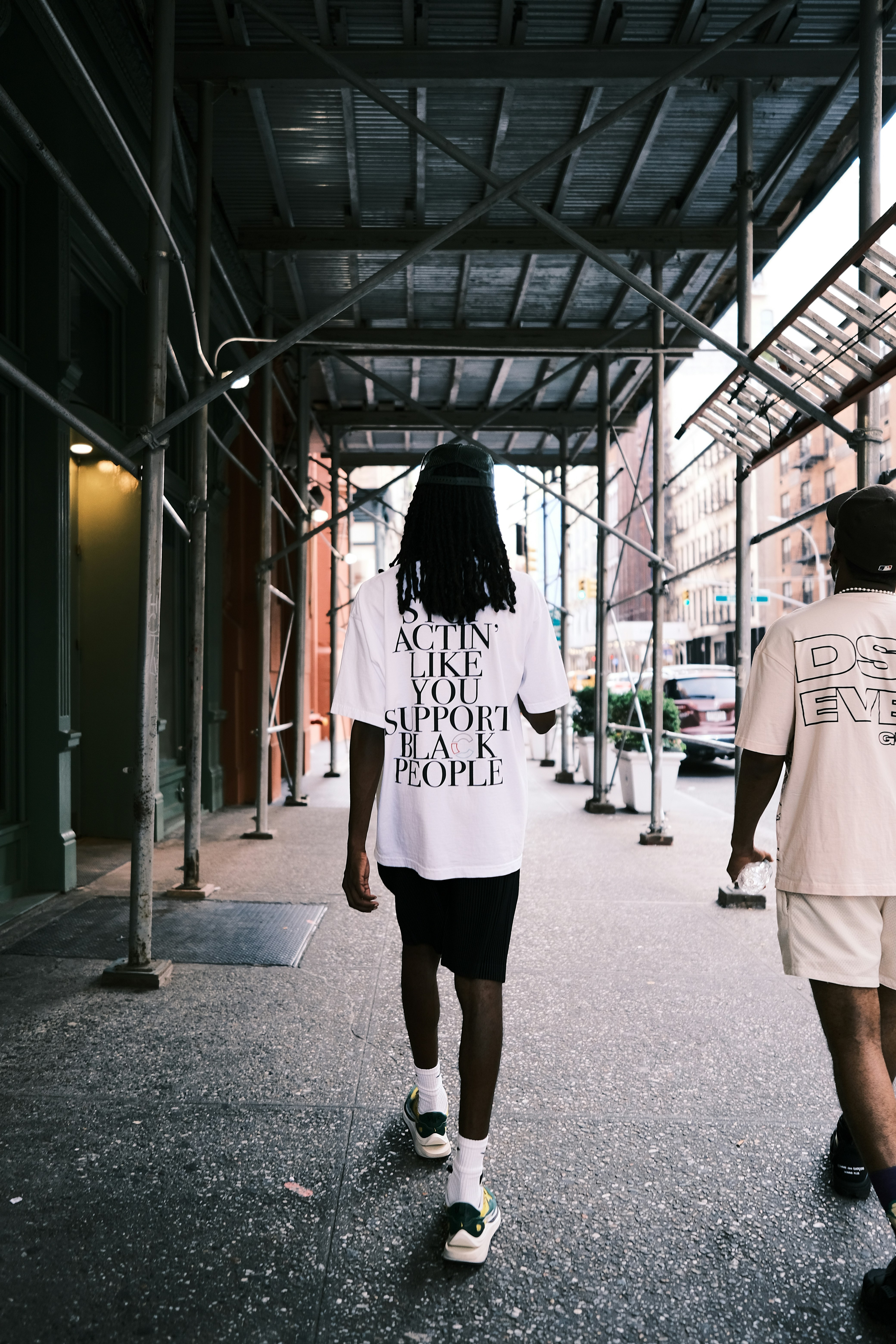 Two individuals walking under a scaffolding, showcasing a message of social awareness on their shirts. The urban setting highlights the blend of fashion and activism.