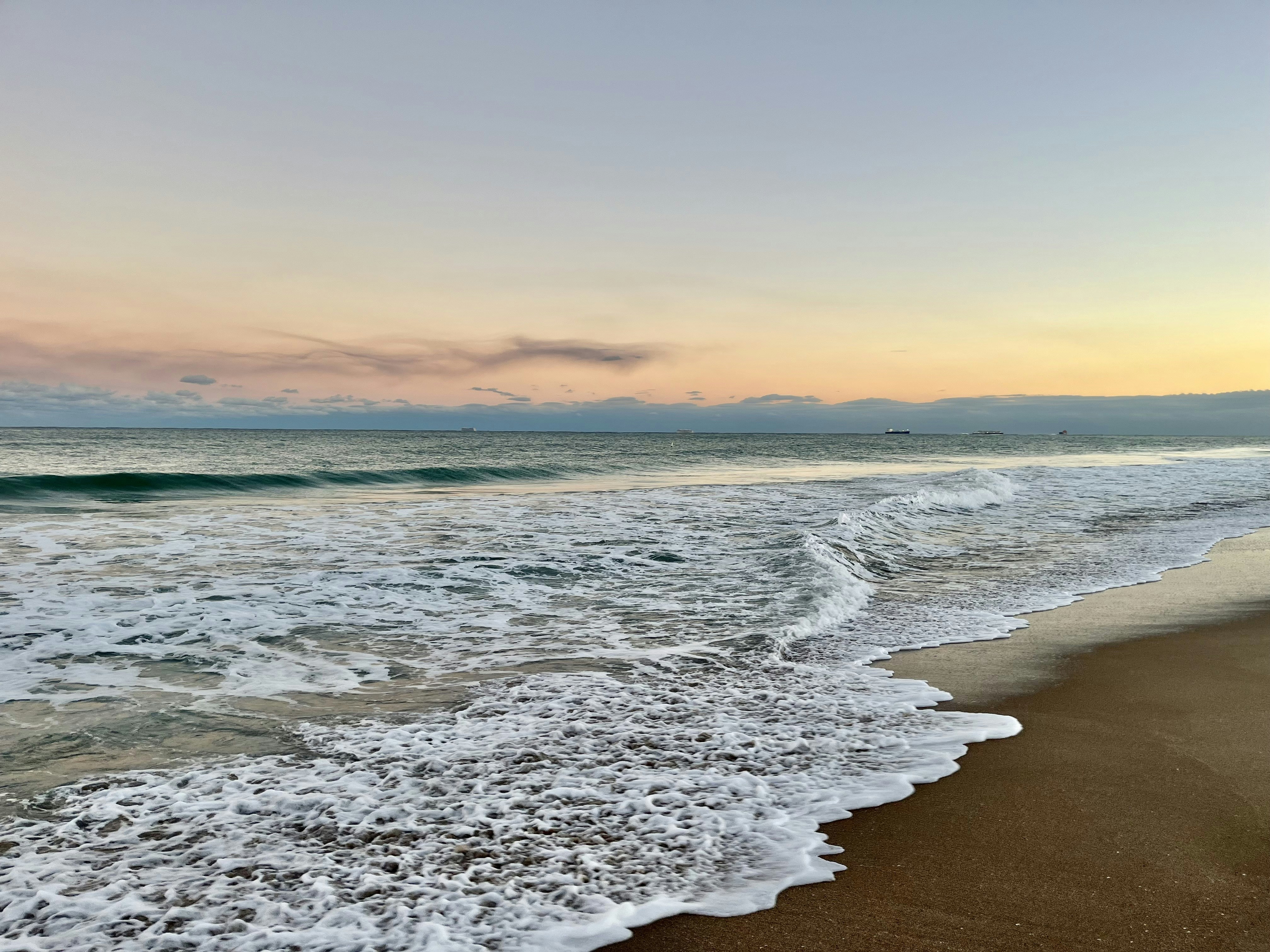 Gentle waves caress a sandy shoreline under a pastel-hued sky at sunrise.