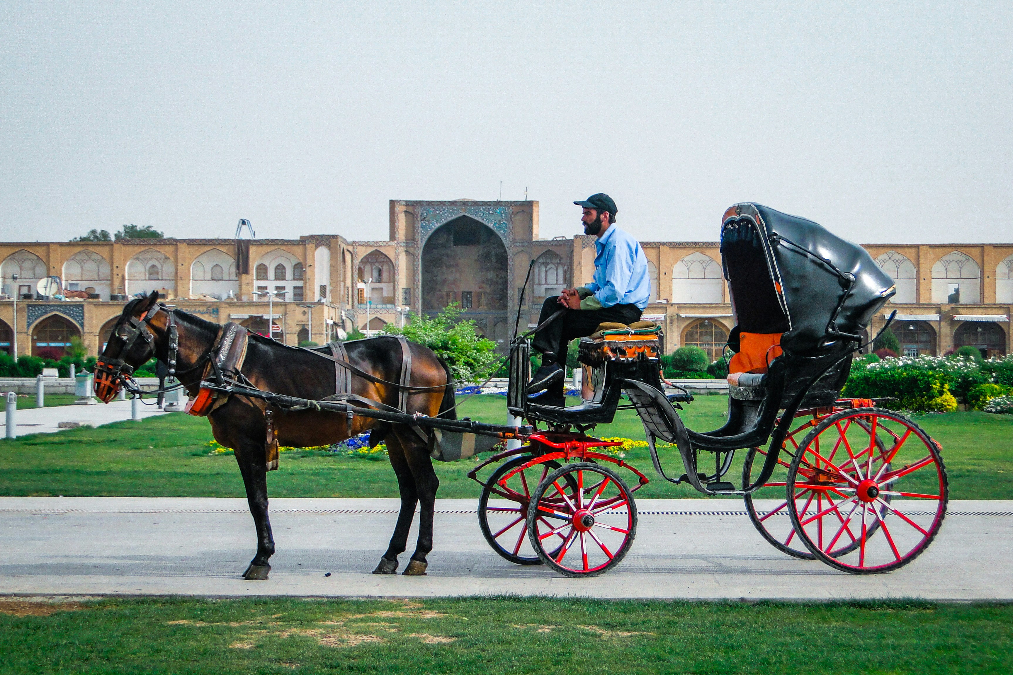 A horse-drawn carriage with a coachman sits in profile beside a historic arched complex, captured on a wide, sunny day. The scene emphasizes the vehicle and its driver against a historic backdrop.
