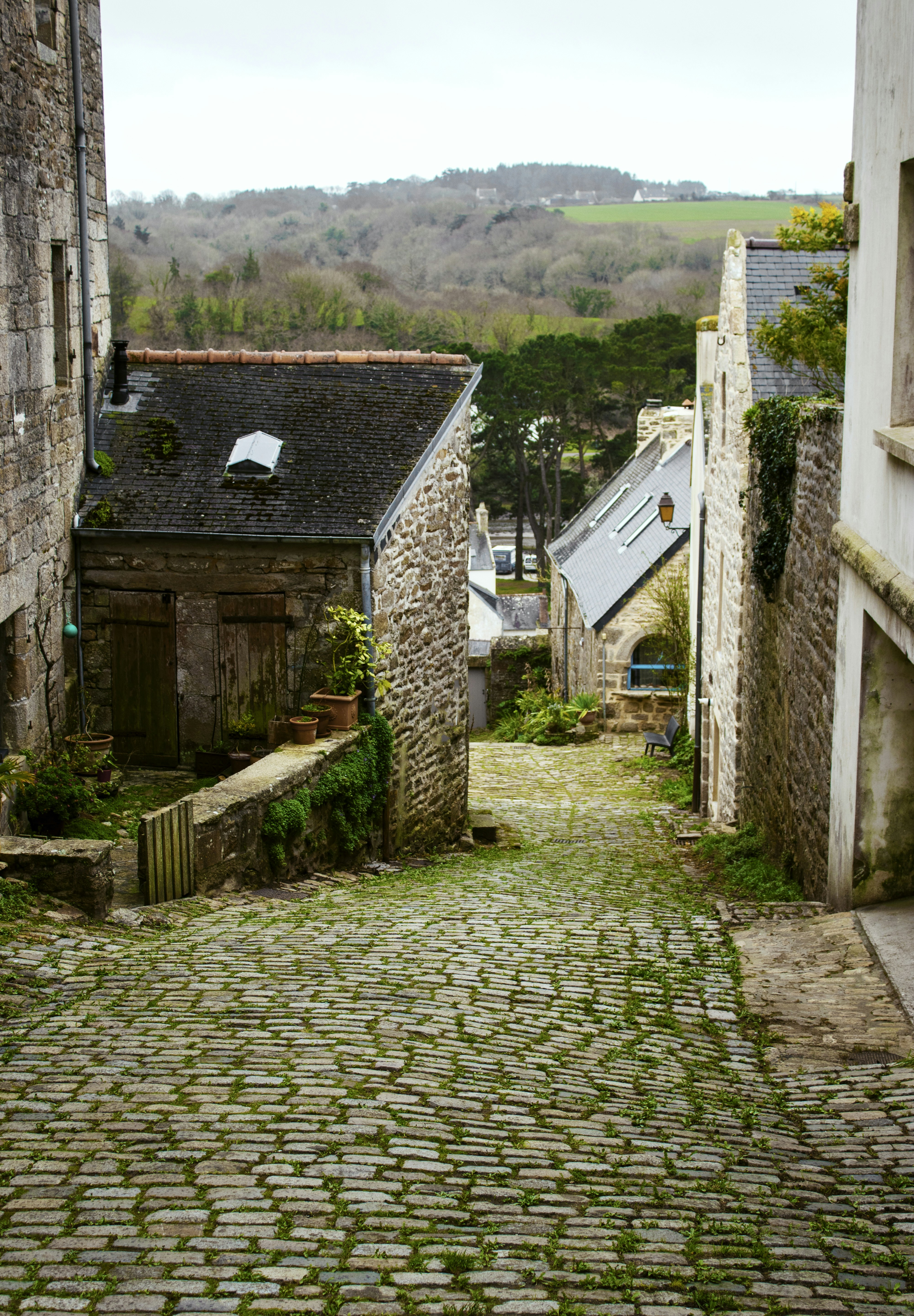 Charming cobblestone alleyway lined with stone buildings and greenery, leading towards a lush landscape in the background.