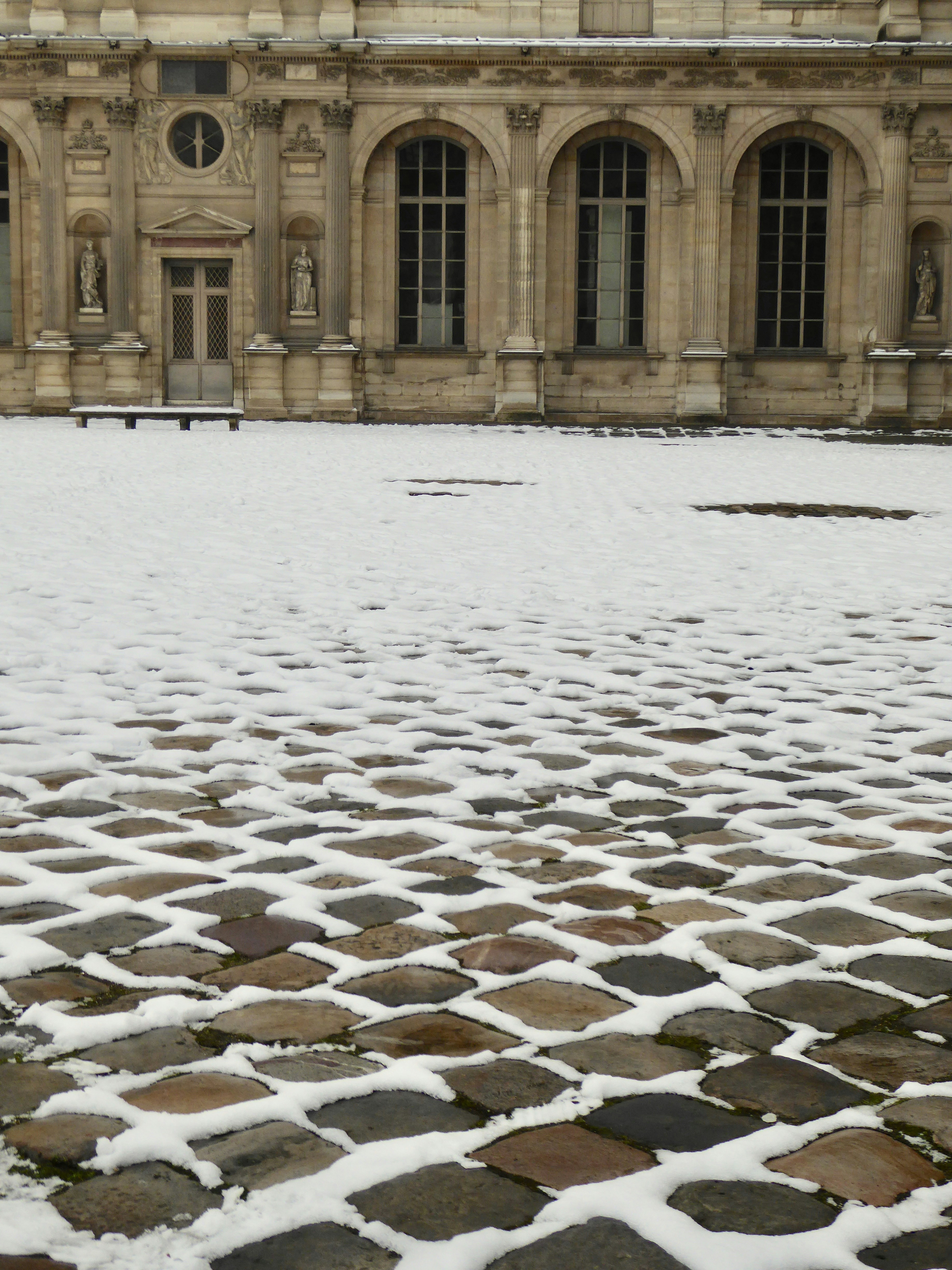 Snow blankets a diamond-patterned cobblestone courtyard before a Beaux-Arts facade with tall arched windows and statues. The scene emphasizes architectural symmetry and a quiet winter mood.