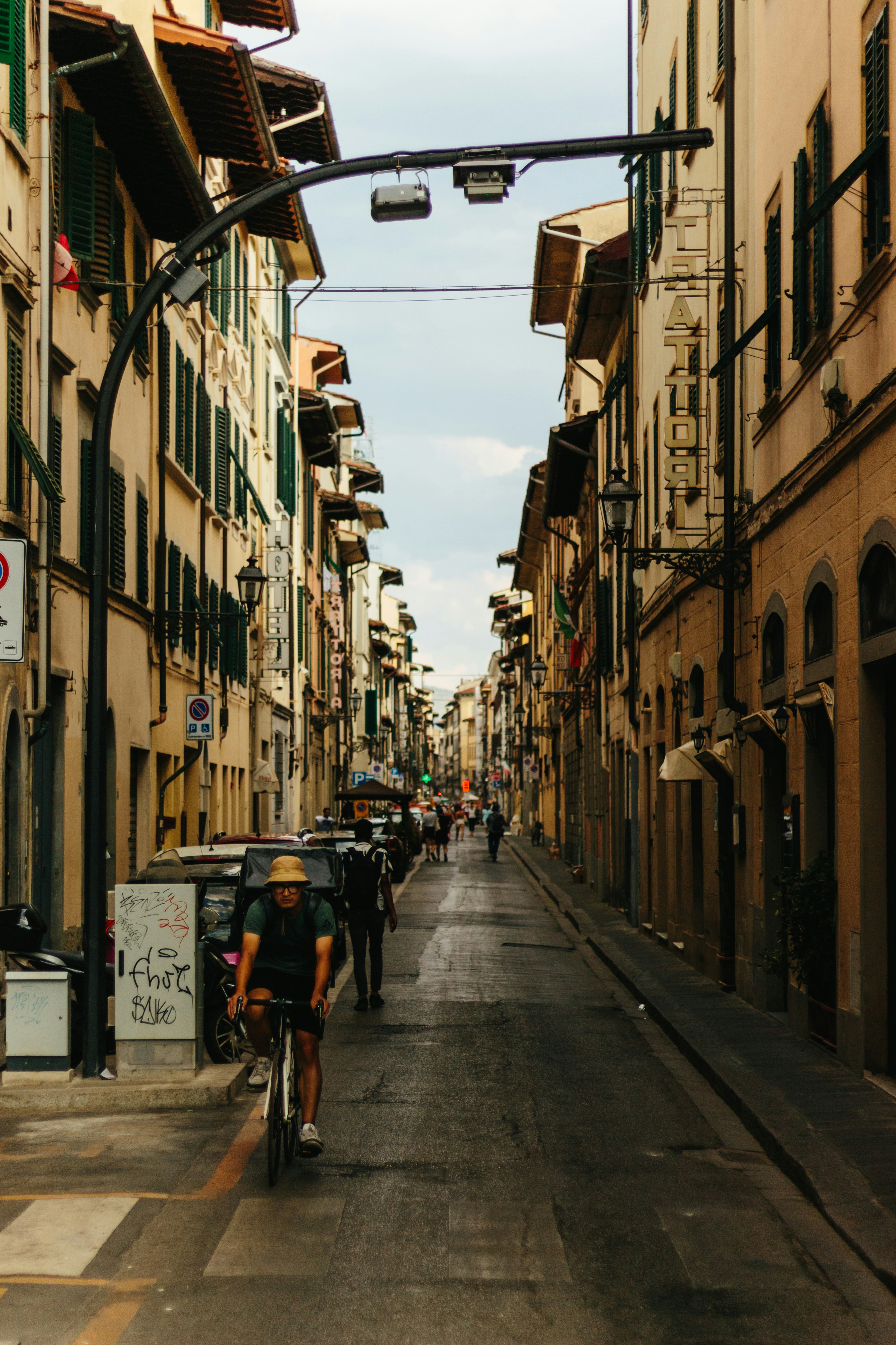 A cyclist navigates a narrow street lined with historic buildings adorned with green shutters in an Italian city. The vibrant atmosphere captures the essence of urban life.