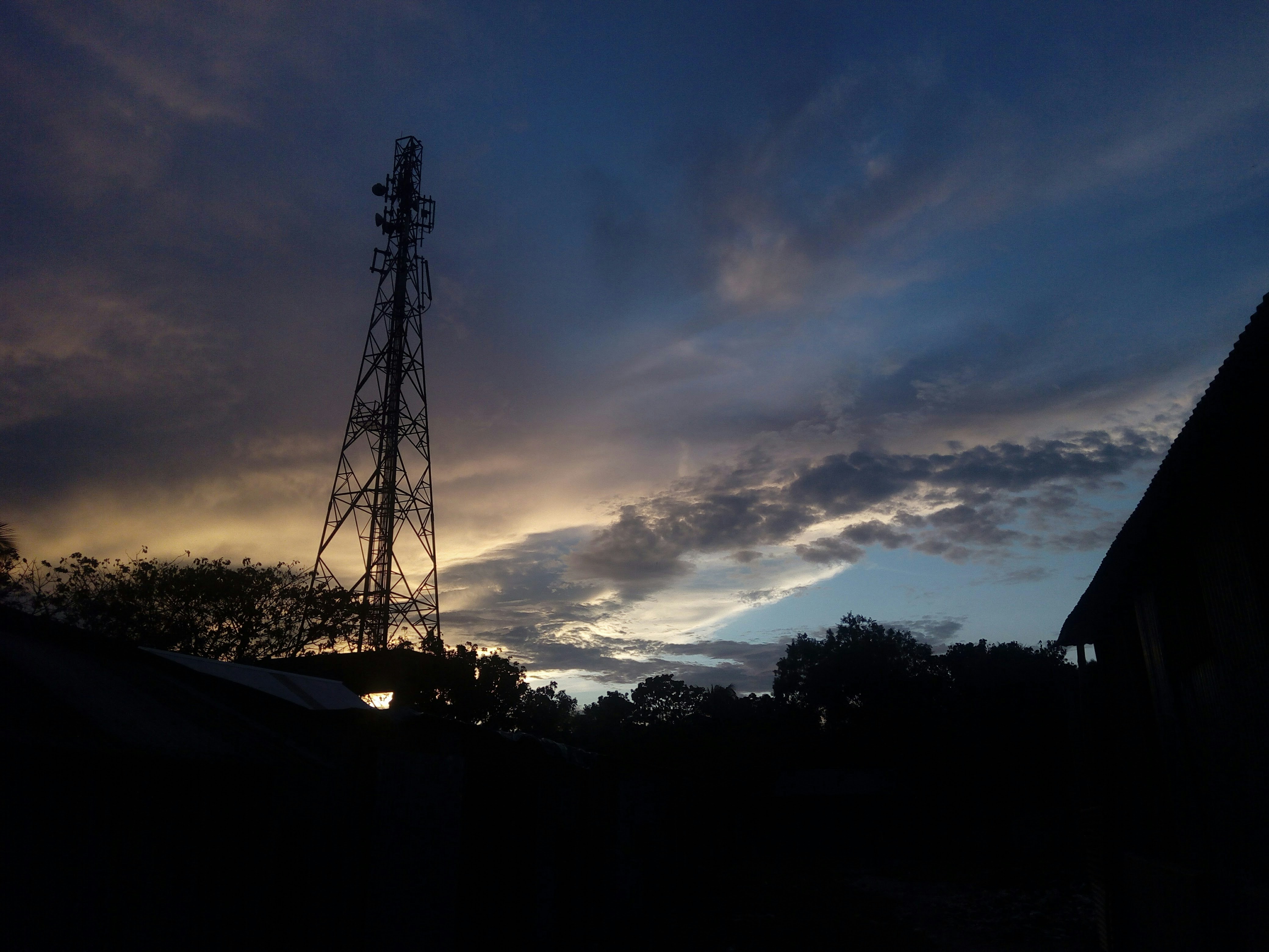 Silhouetted telecommunications tower against a dramatic twilight sky, framed by darkened foliage and buildings. The scene captures the transition from day to night.