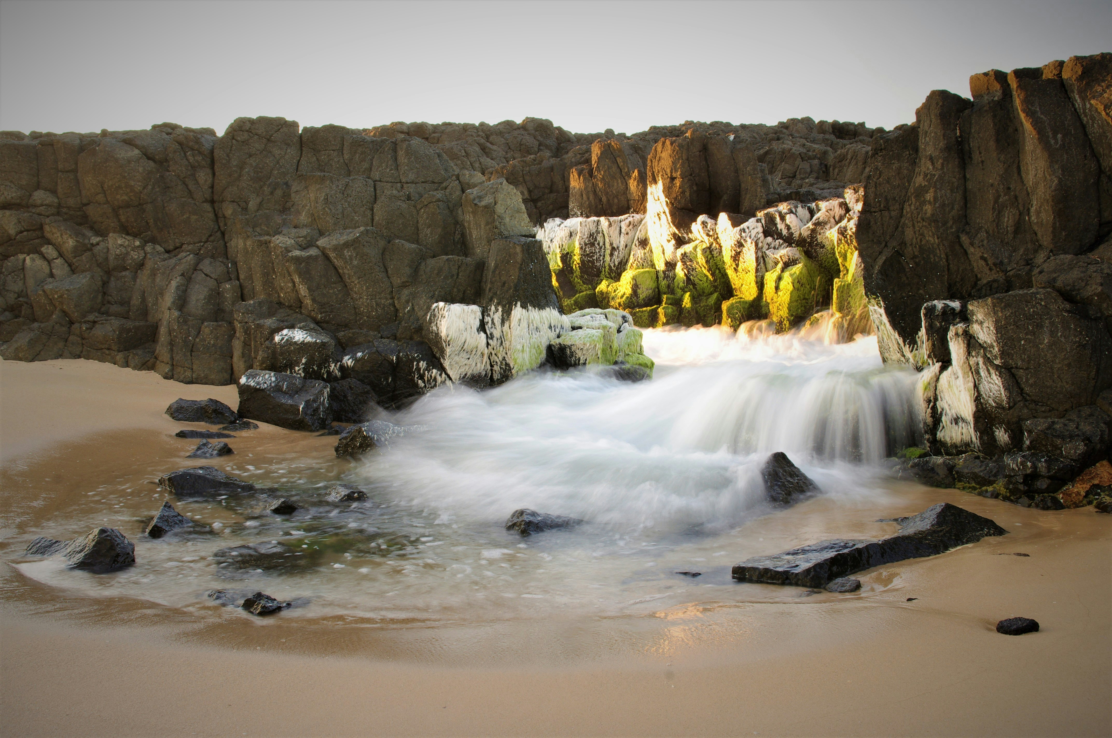 Water flows gently through rocky formations, with vibrant moss contrasting against the rugged stones. The soft sand completes the tranquil coastal scene.