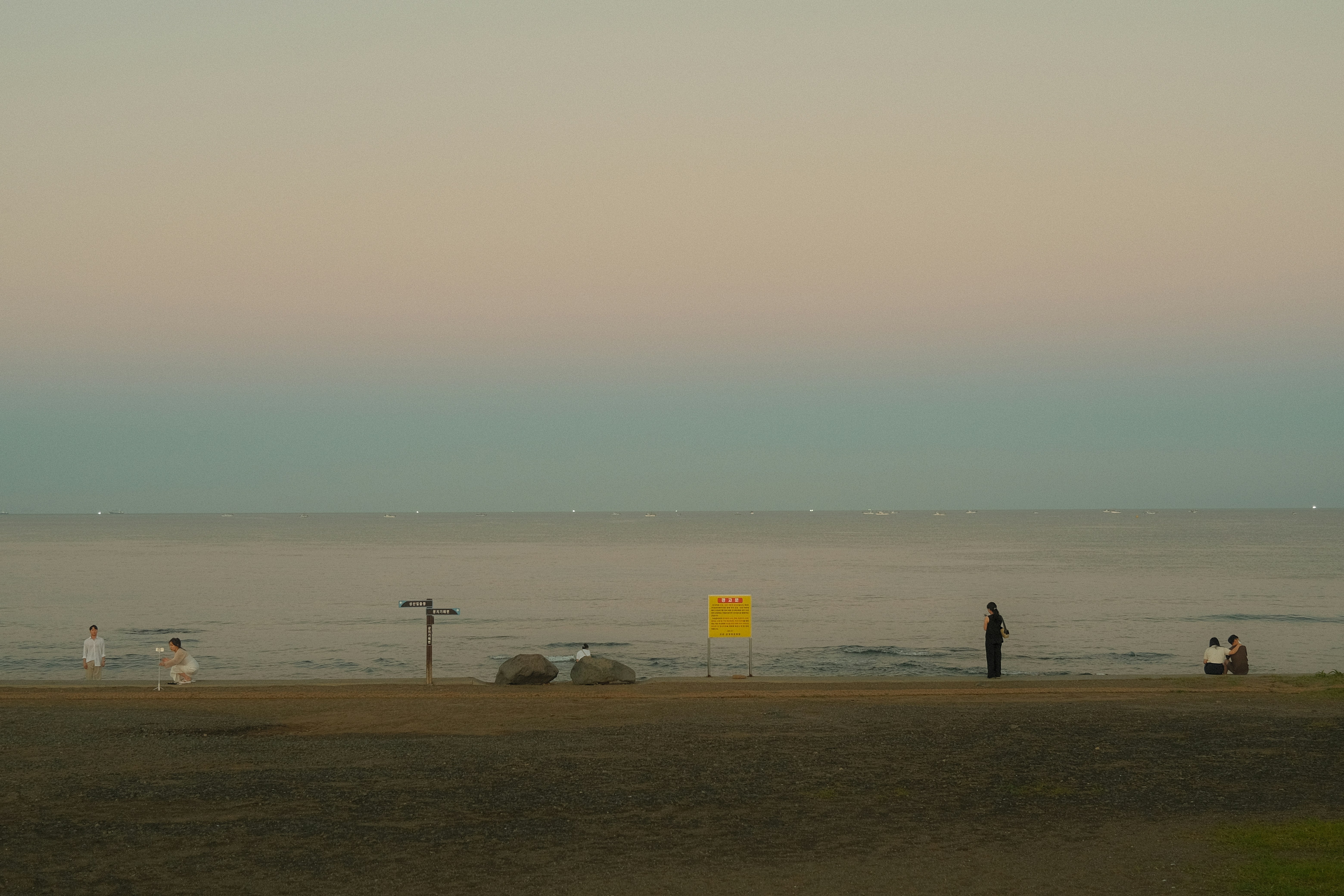 A serene beach scene at dusk, featuring figures along the shoreline and a warning sign. The soft gradient of the sky transitions from pastel hues to a calm sea.