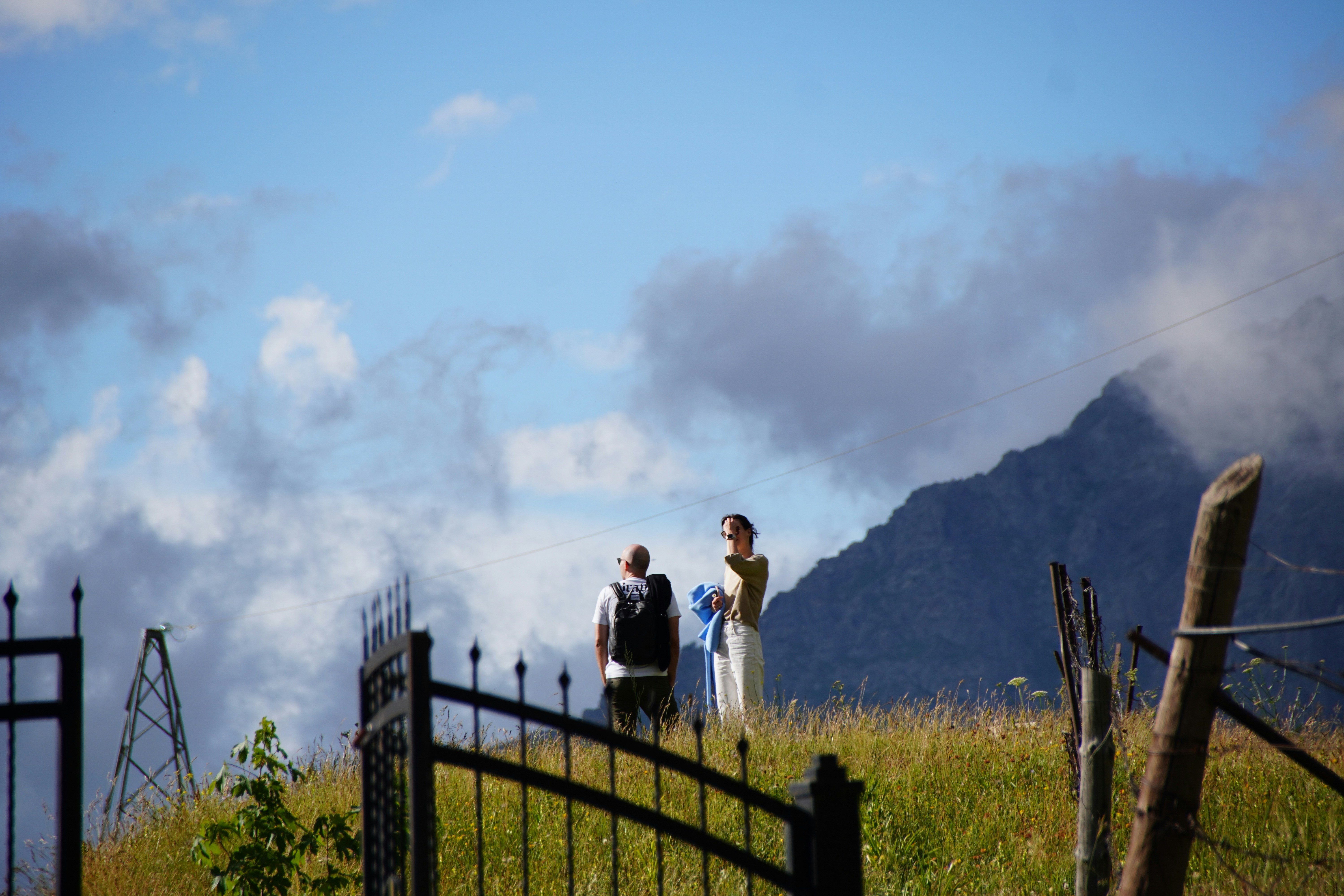 Two hikers on a sunlit ridge framed by a weathered fence and distant Alpine peak