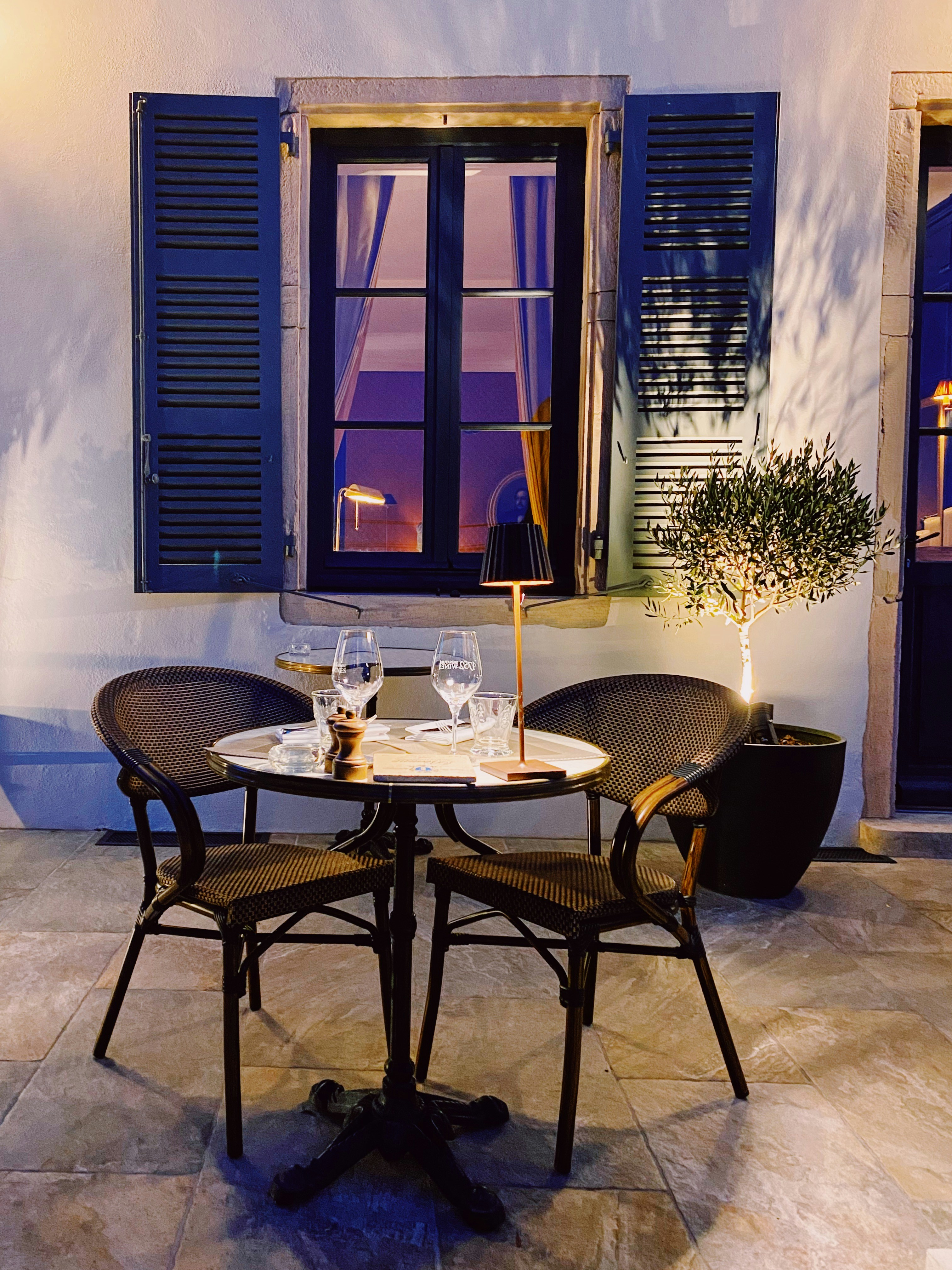 A charming outdoor dining setup featuring a round table with wine glasses and a small lamp, framed by blue shutters and a potted plant. The warm evening light adds a cozy atmosphere.