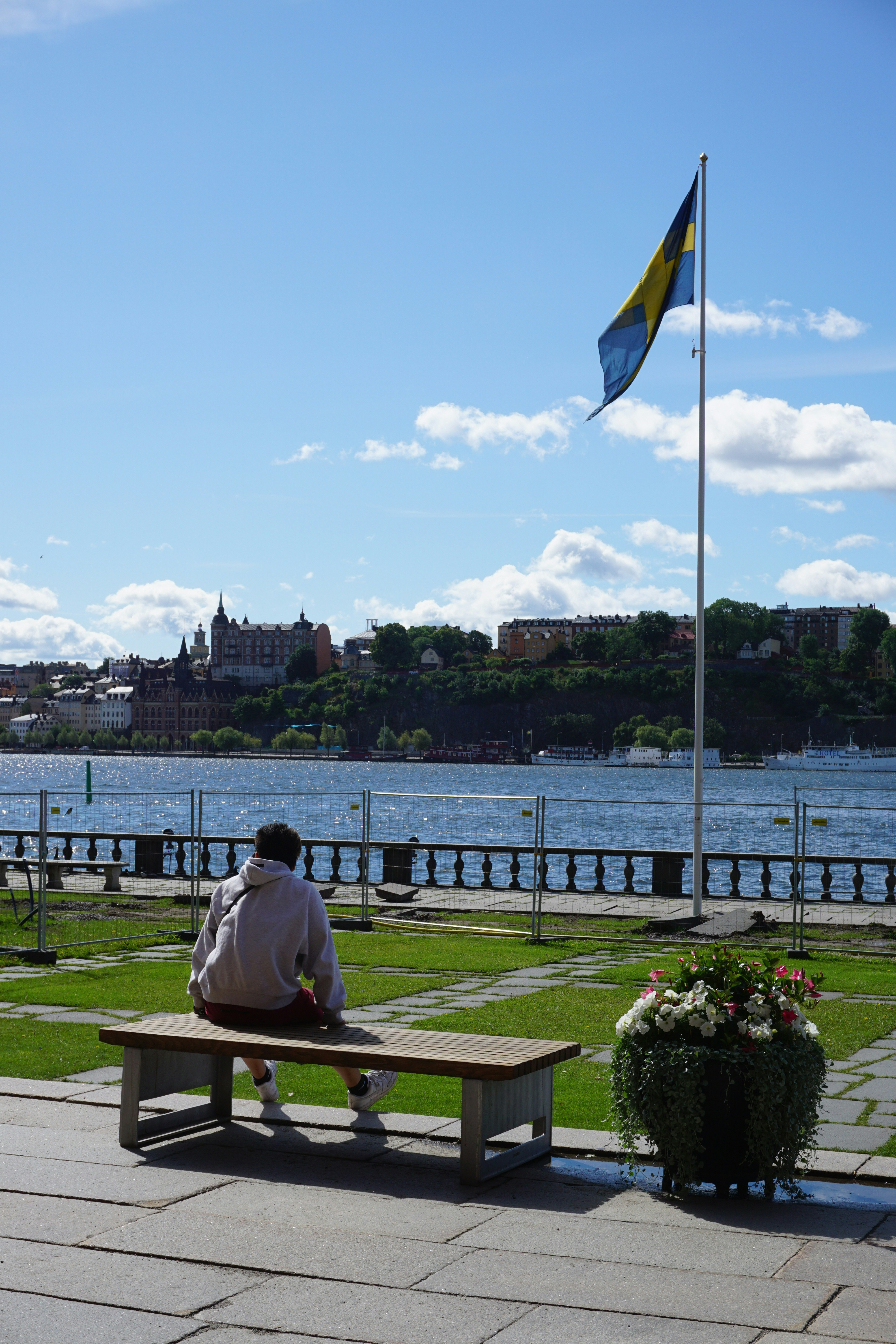 A person sits on a bench, gazing at the waterfront view adorned with a flag and lush greenery. The scene captures a tranquil atmosphere by the riverside.