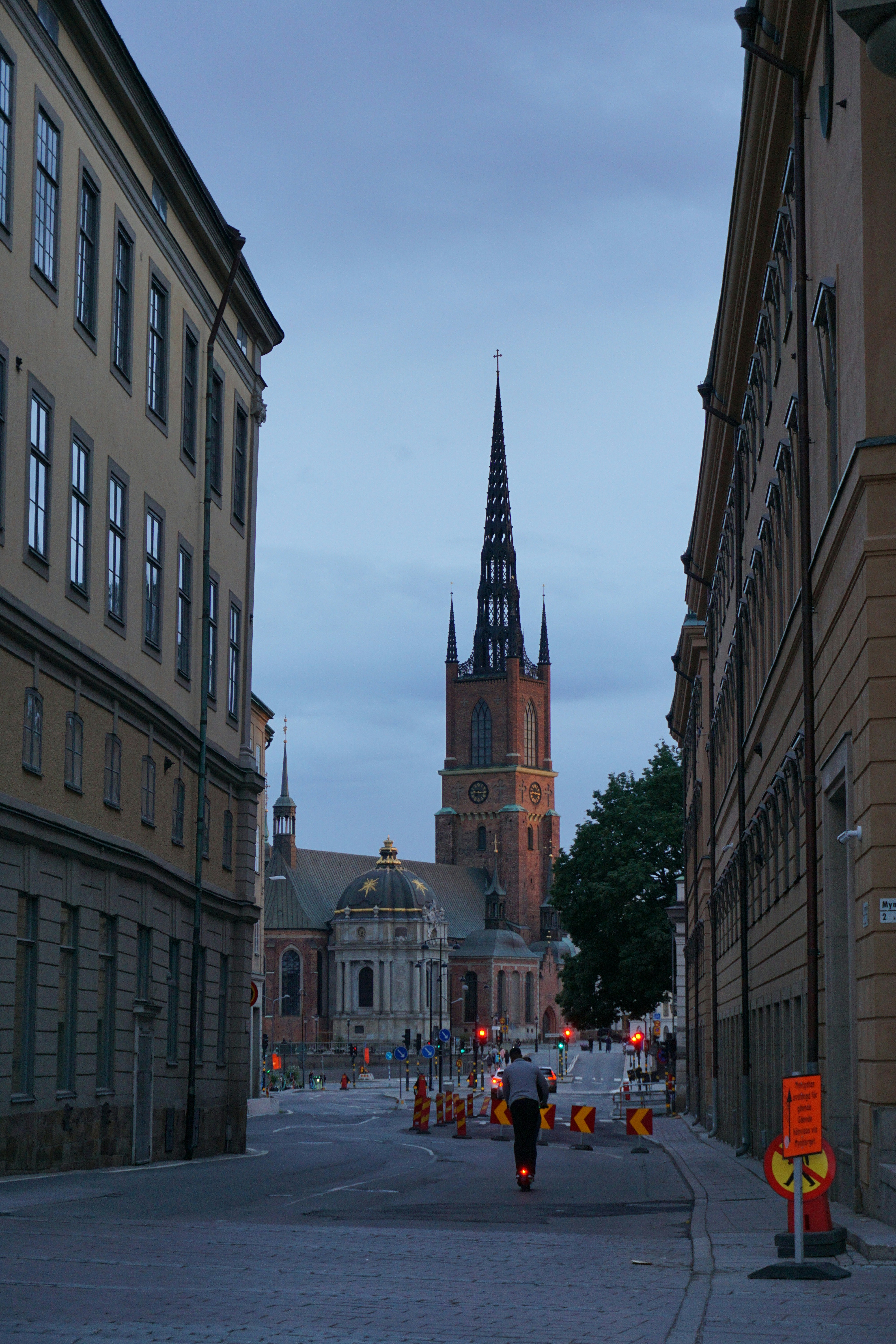 A narrow street framed by historic buildings leads to a tall church spire, illuminated in the evening light. The scene captures the tranquil ambiance of an urban landscape at dusk.