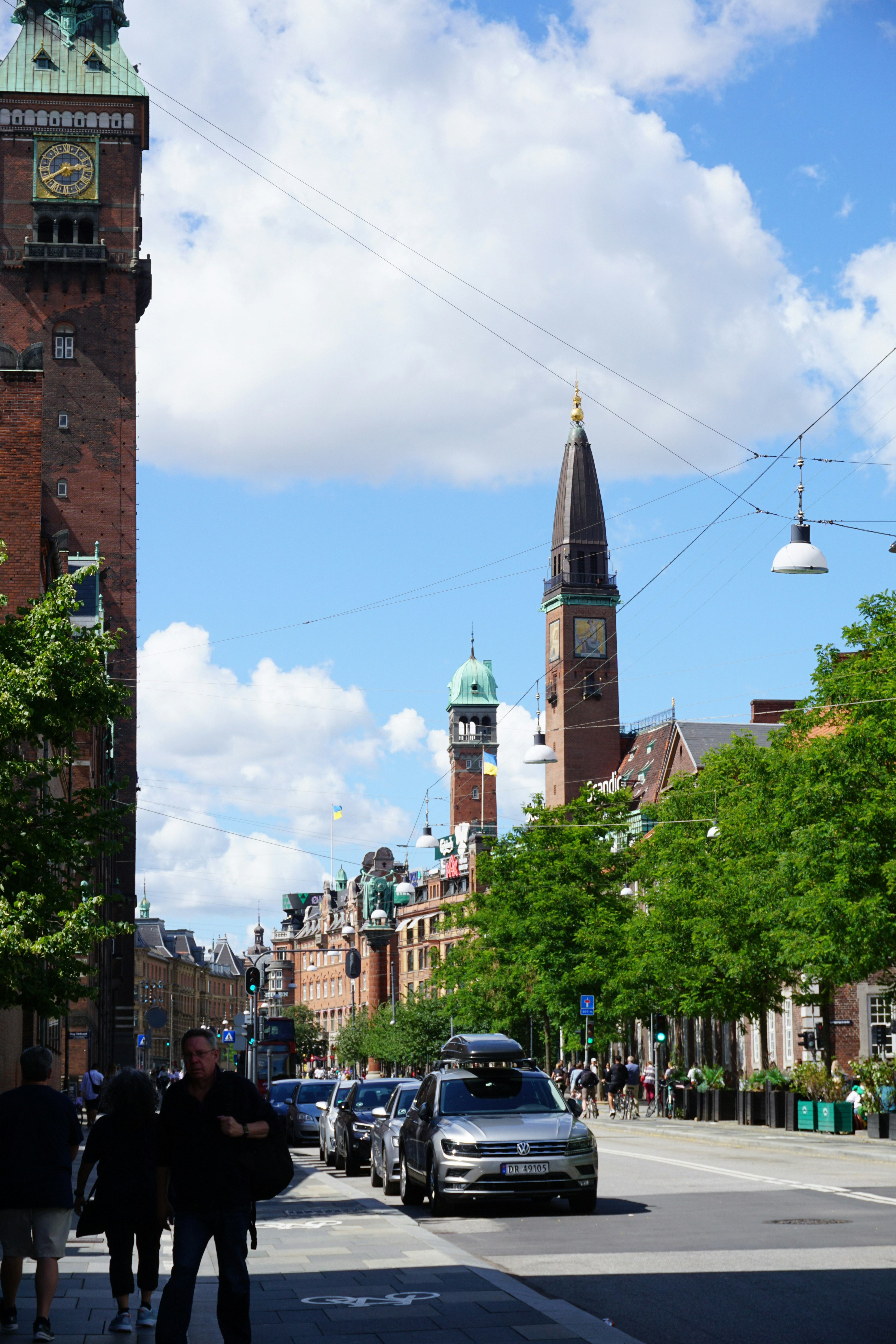 Historical buildings line a bustling street in Copenhagen, framed by vibrant trees and a bright blue sky. The scene captures the lively atmosphere of the city.