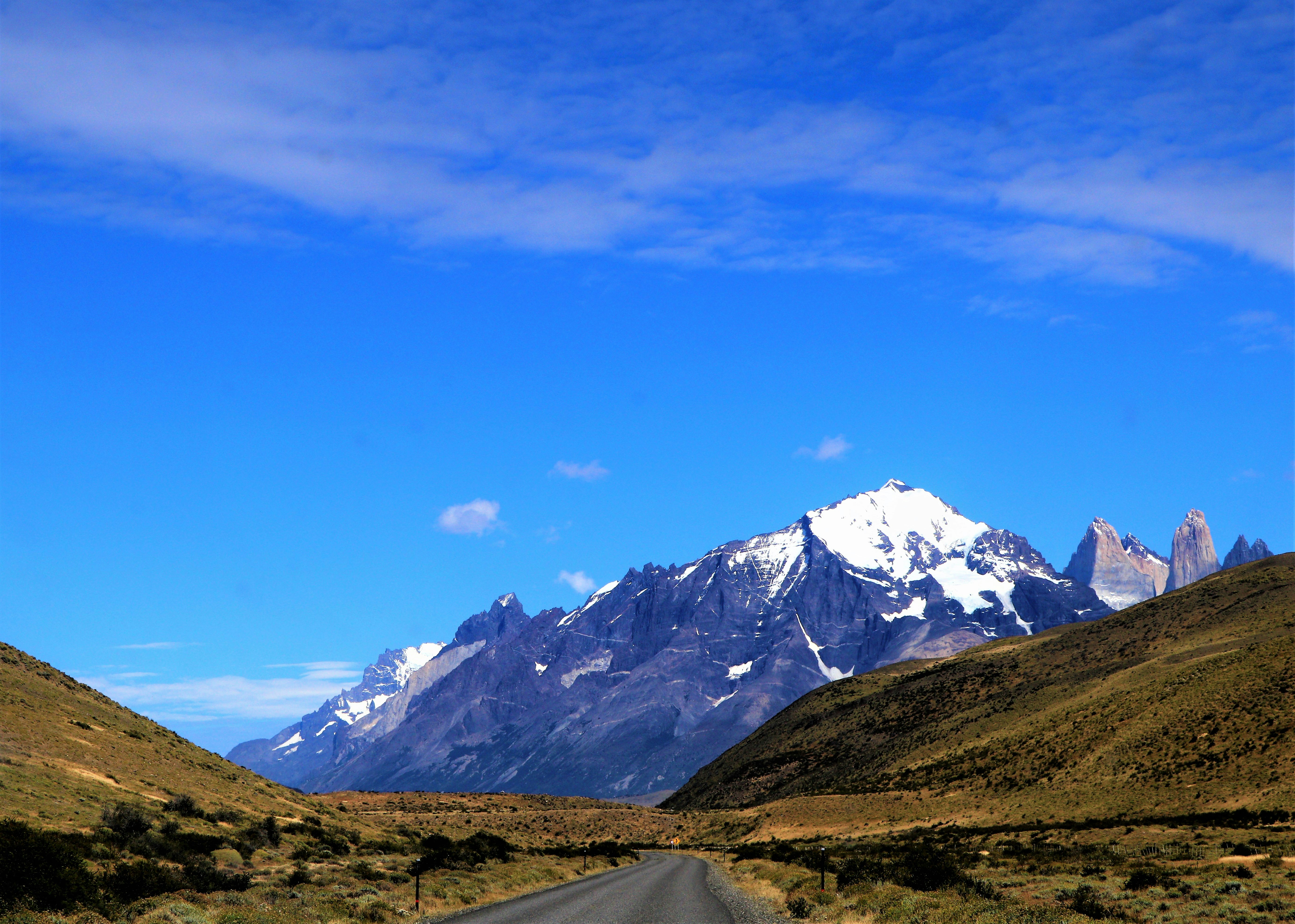 This breathtaking image captures a serene road leading towards snow-capped mountains, set against a vivid blue sky. The composition beautifully balances the rugged terrain with the expansive sky, while the lighting highlights the contrast between the dark mountains and their gleaming snowcaps. The rich blues and earthy tones create a striking visual contrast, enhancing the grandeur and tranquility of the landscape.