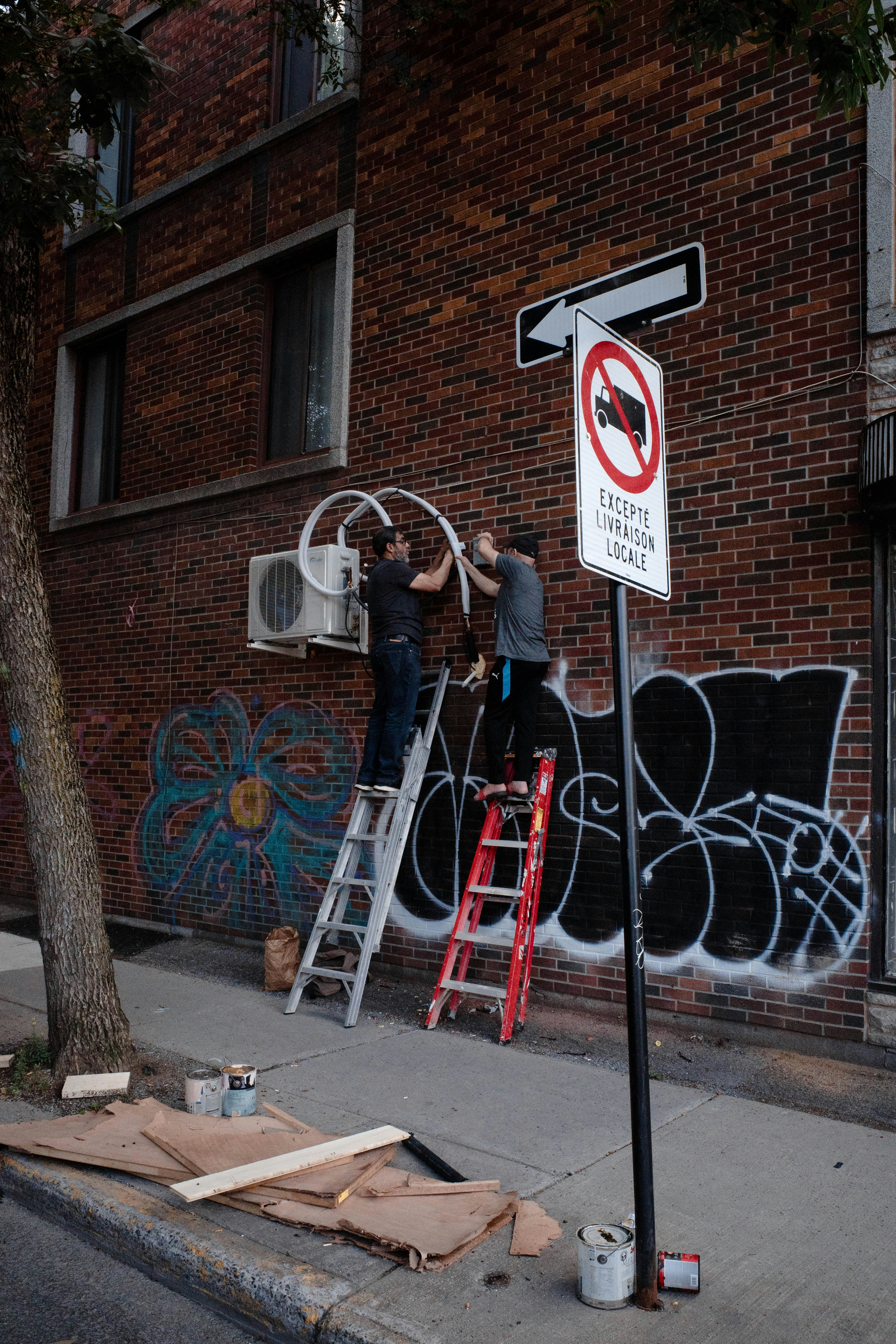 Urban Fix: Two Ladder Workers Secure a Circular HVAC Ring on Graffiti-Scrawled Brick