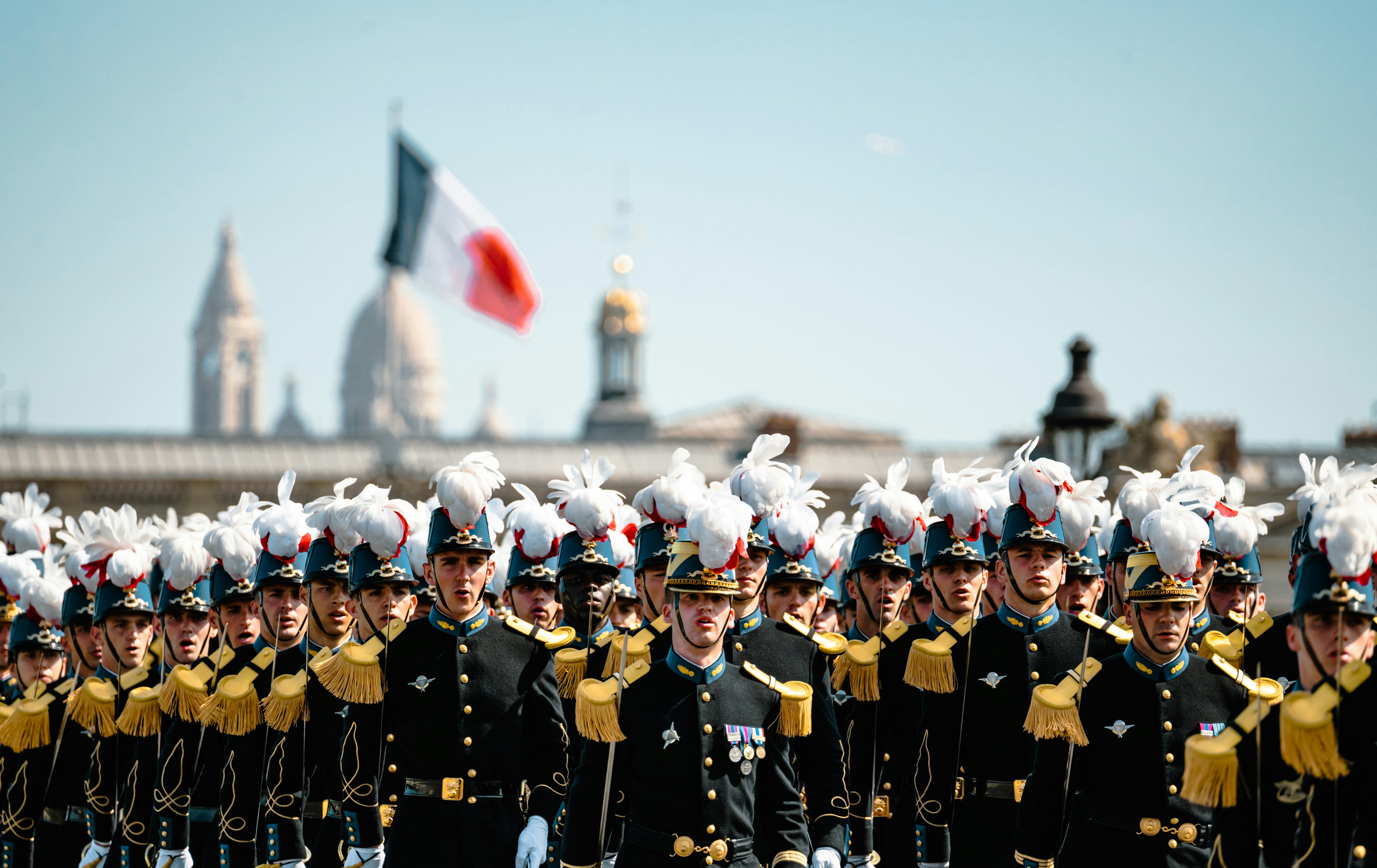 Uniformed soldiers parade in formation, showcasing their ceremonial attire against a backdrop of historic architecture and the French flag. 