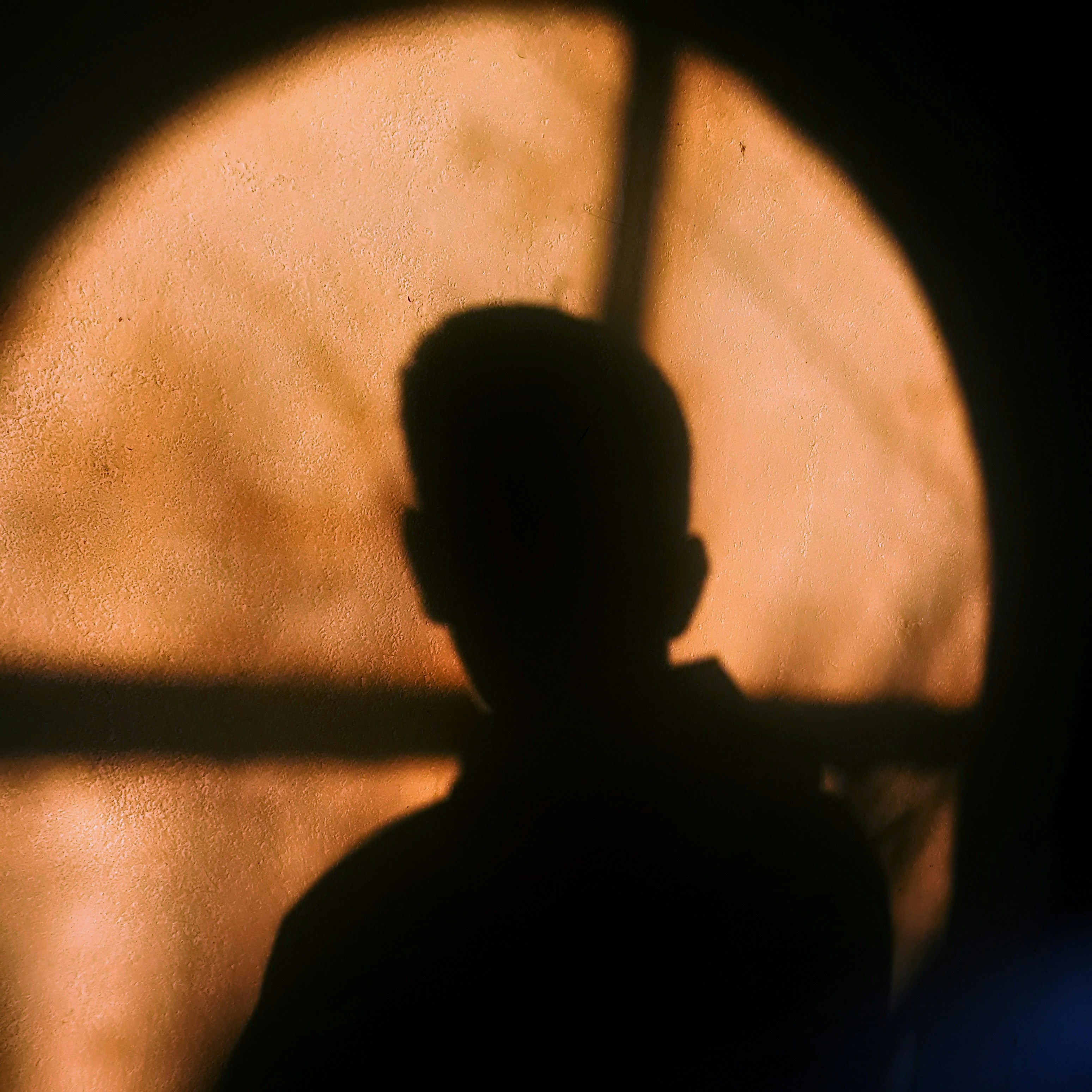 A shadowy figure stands in front of a large, softly illuminated backdrop, creating a striking contrast. The warm tones evoke a sense of mystery.