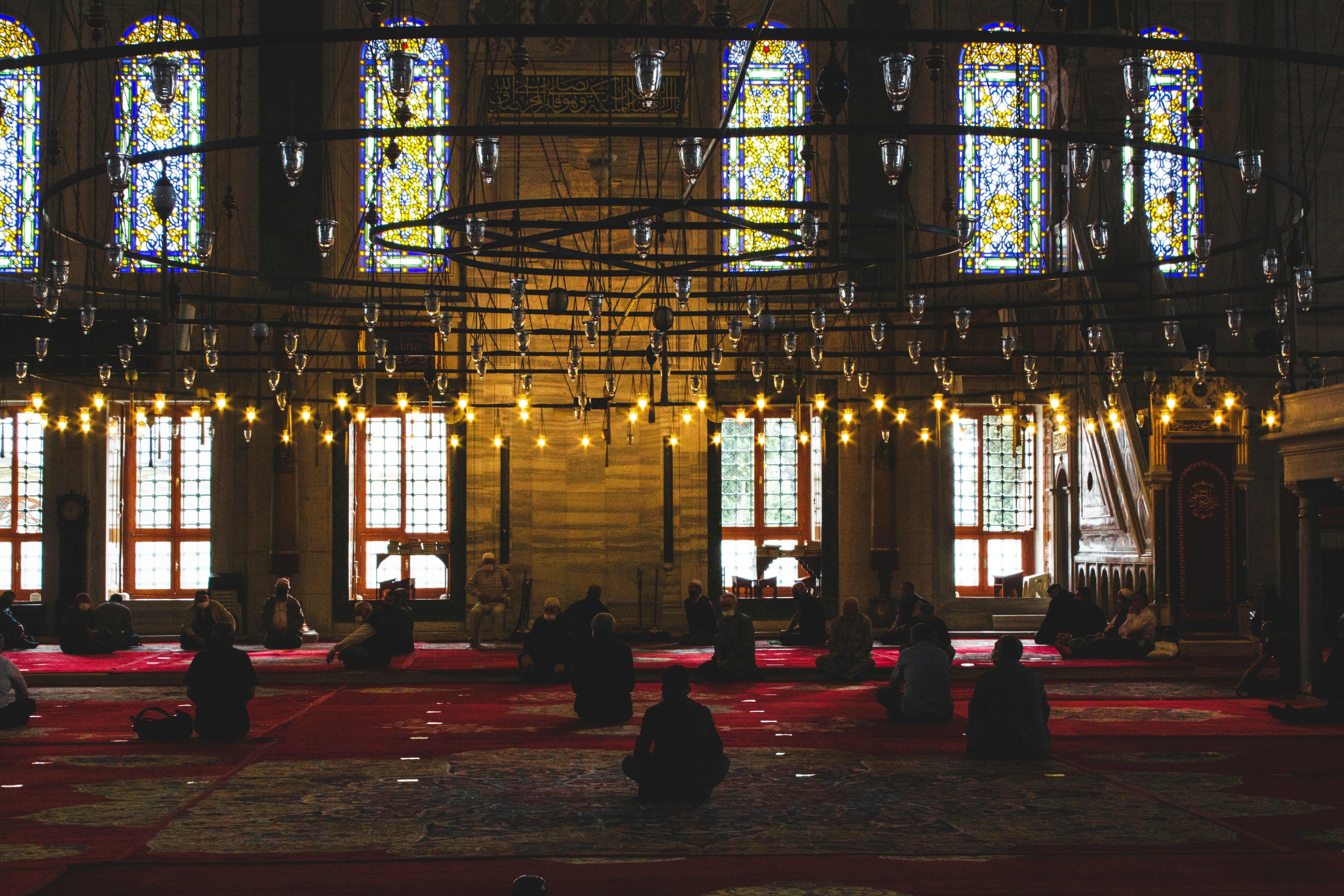 Interior of a mosque featuring intricate stained glass windows and soft lighting, with worshippers seated on ornate carpets.