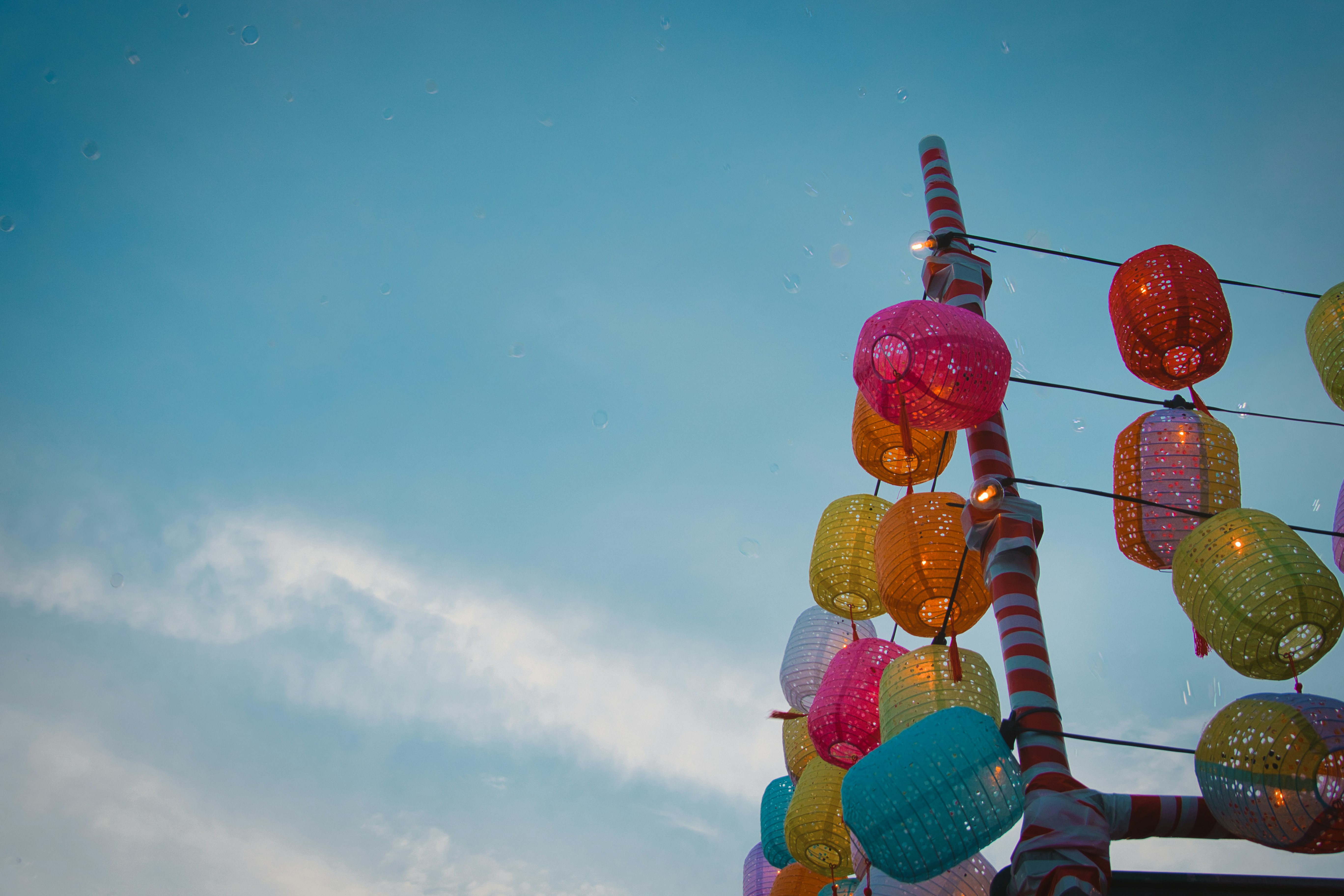 Colorful lanterns hanging from a structure, illuminated against a backdrop of a soft blue sky with wispy clouds.