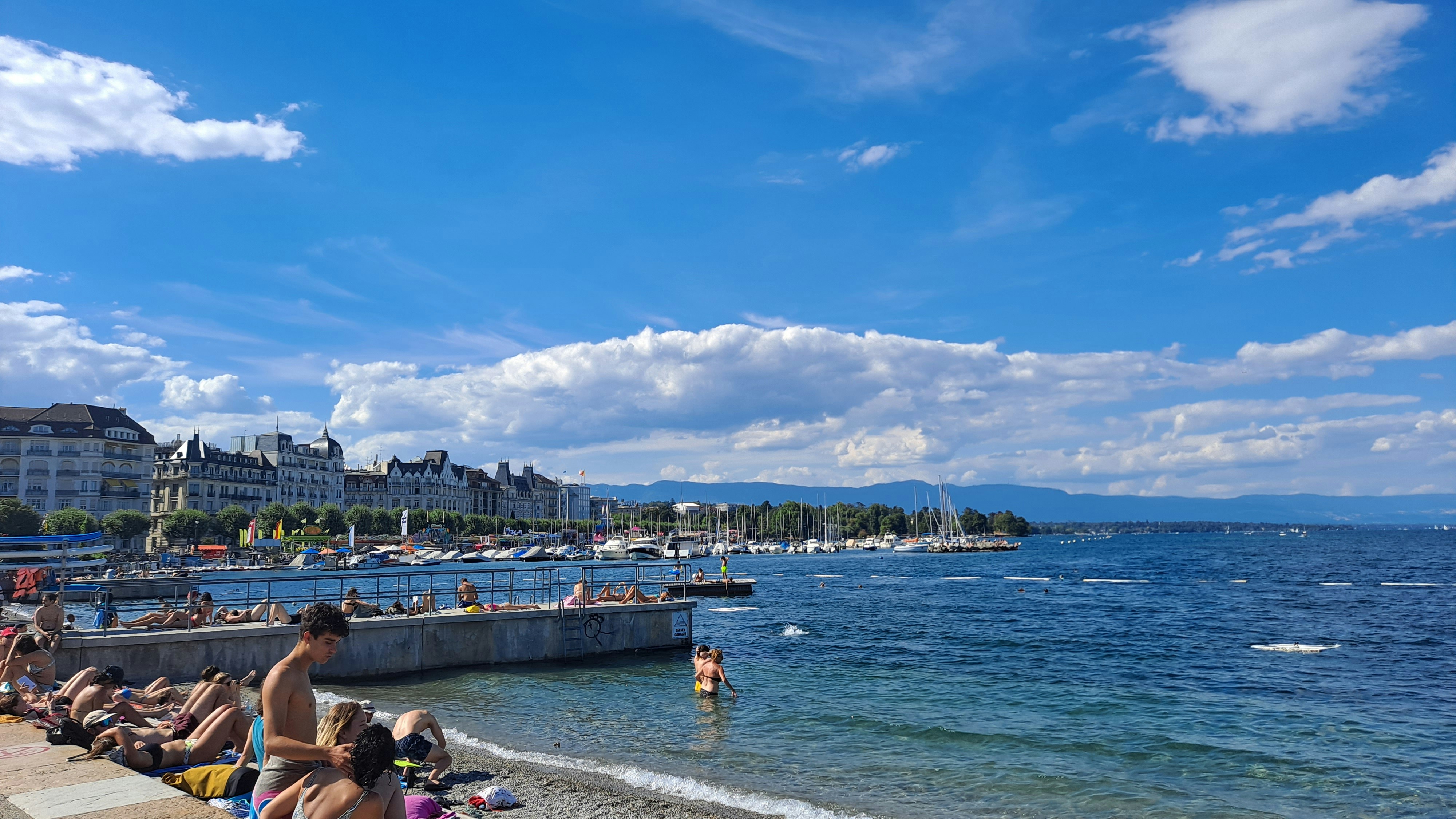 Harborfront in Light: Sunlit Promenade and Sailboats on a Calm Sea