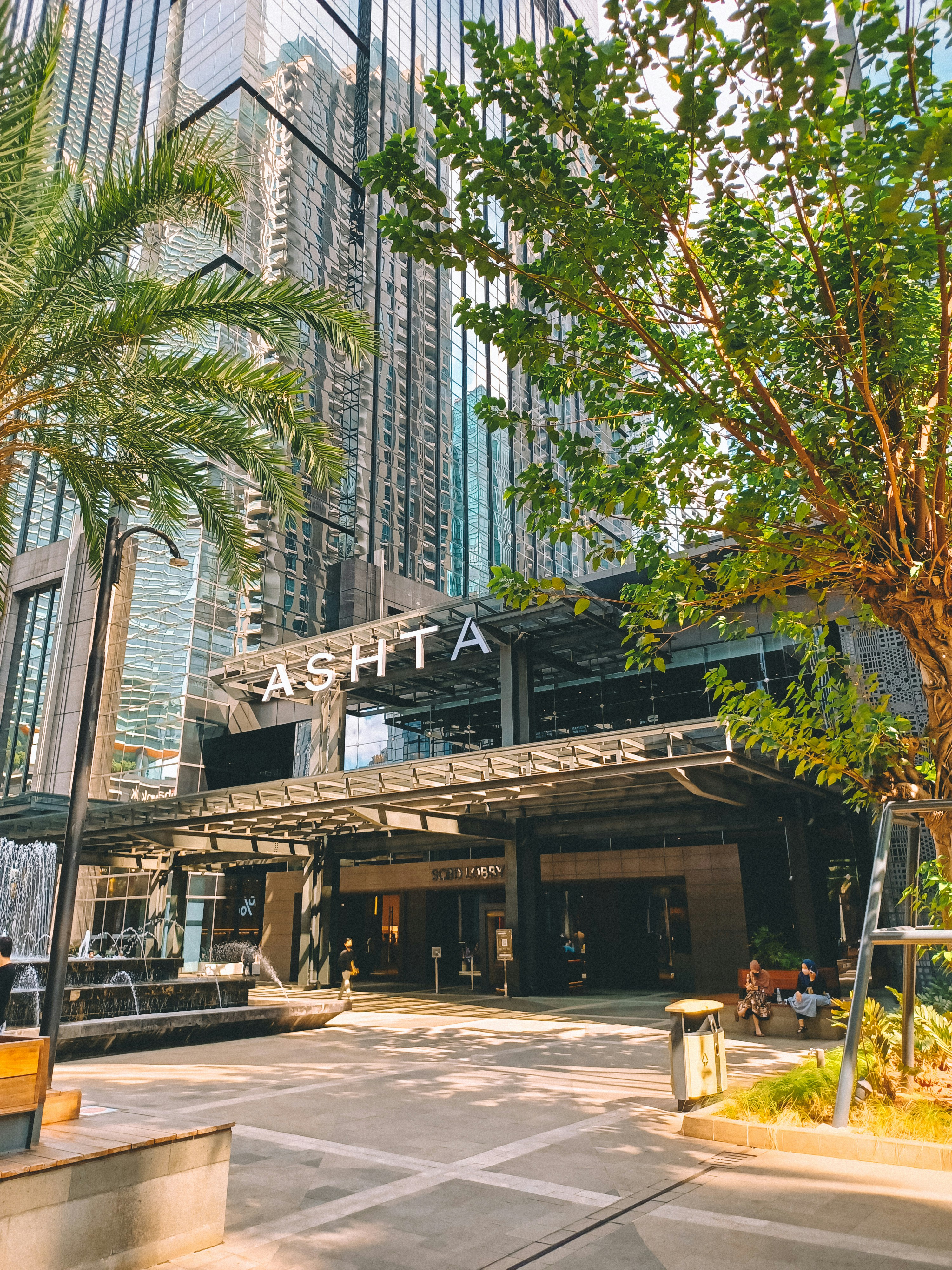 Modern building facade with palm trees and a lively entrance under a clear sky.