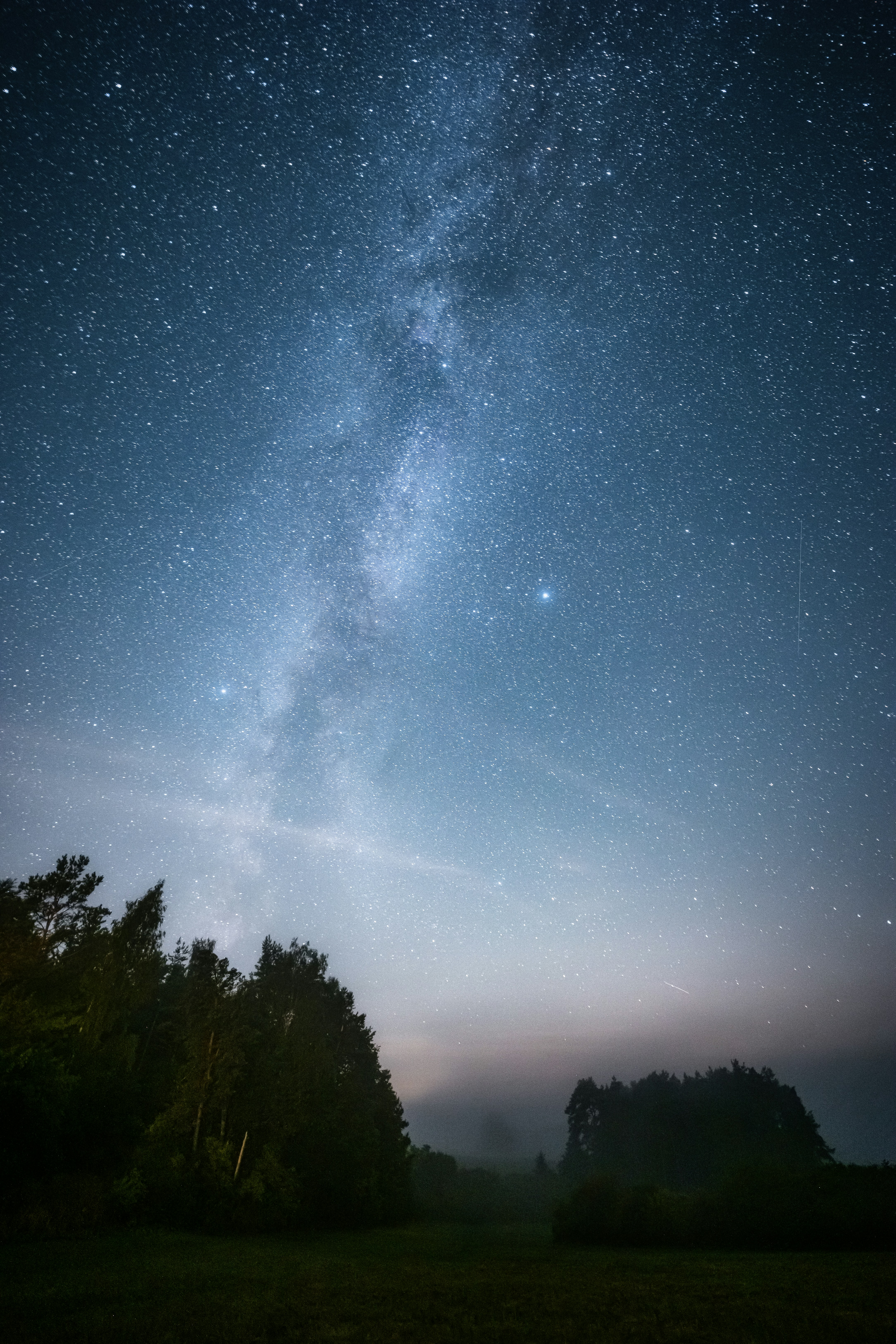 Milky Way stretching across a starry night sky above a peaceful forest landscape.