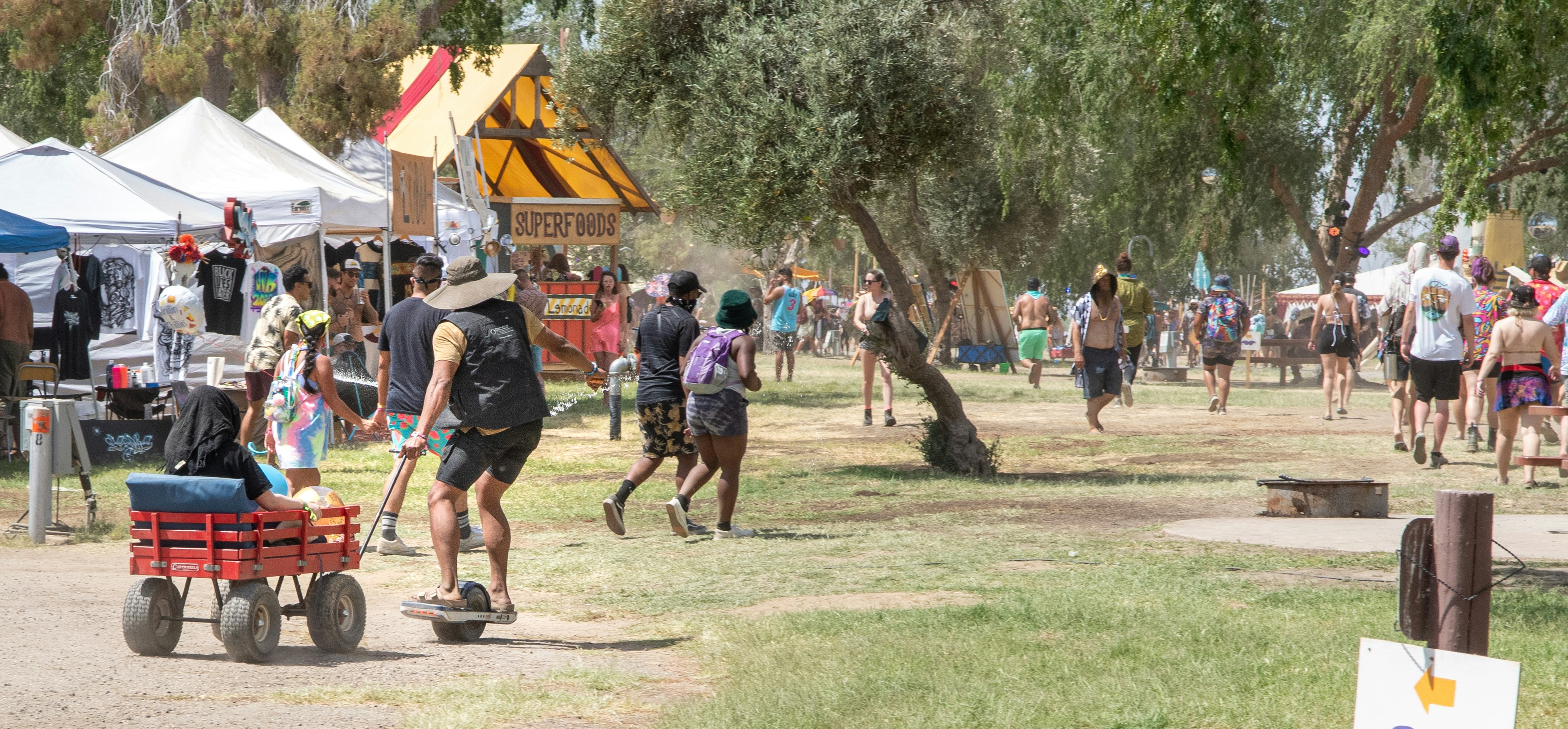 People enjoying an outdoor market with tents and trees under a sunny sky.