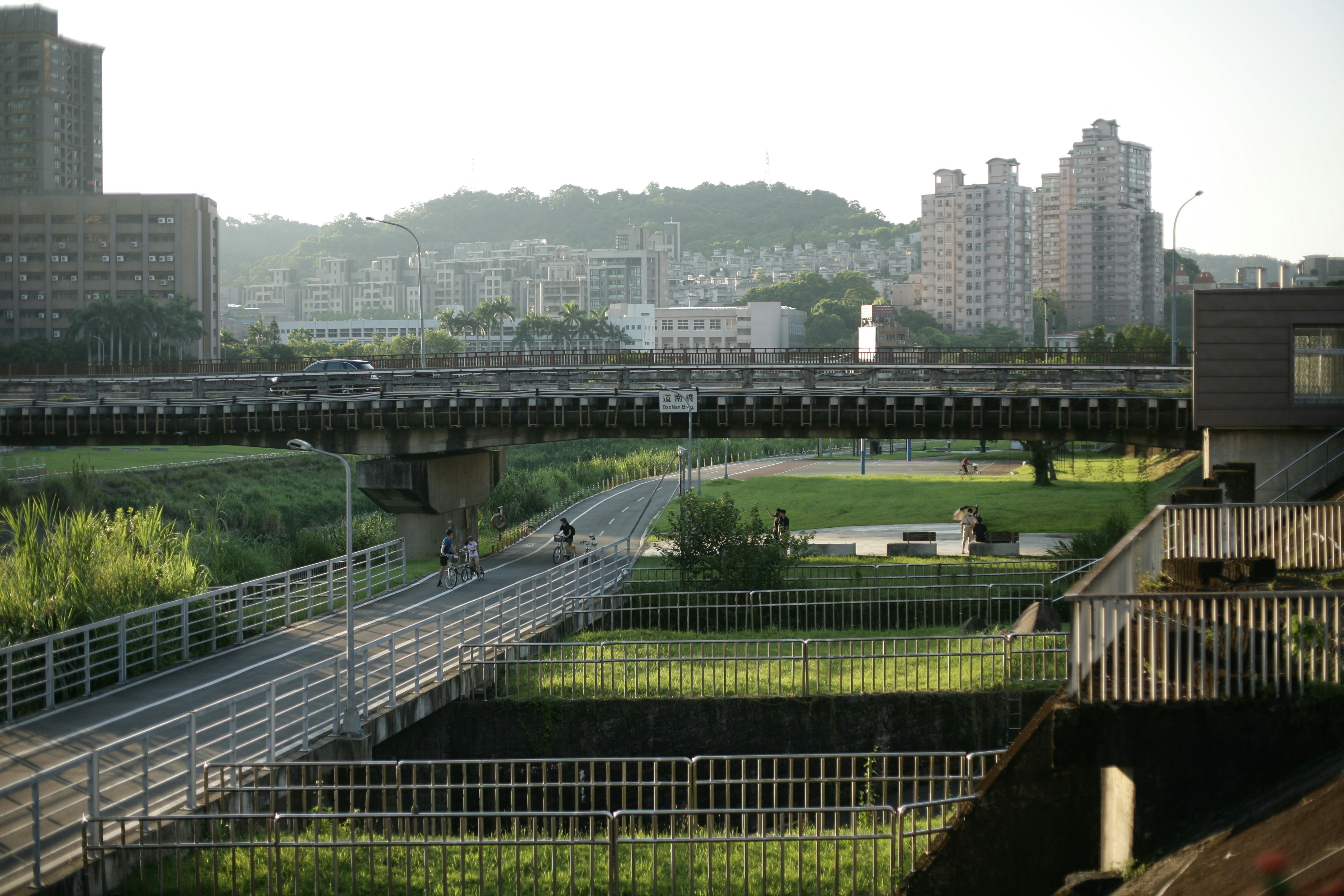 Bicyclists traverse a winding path beneath a bridge, framed by lush greenery and urban buildings in the background.