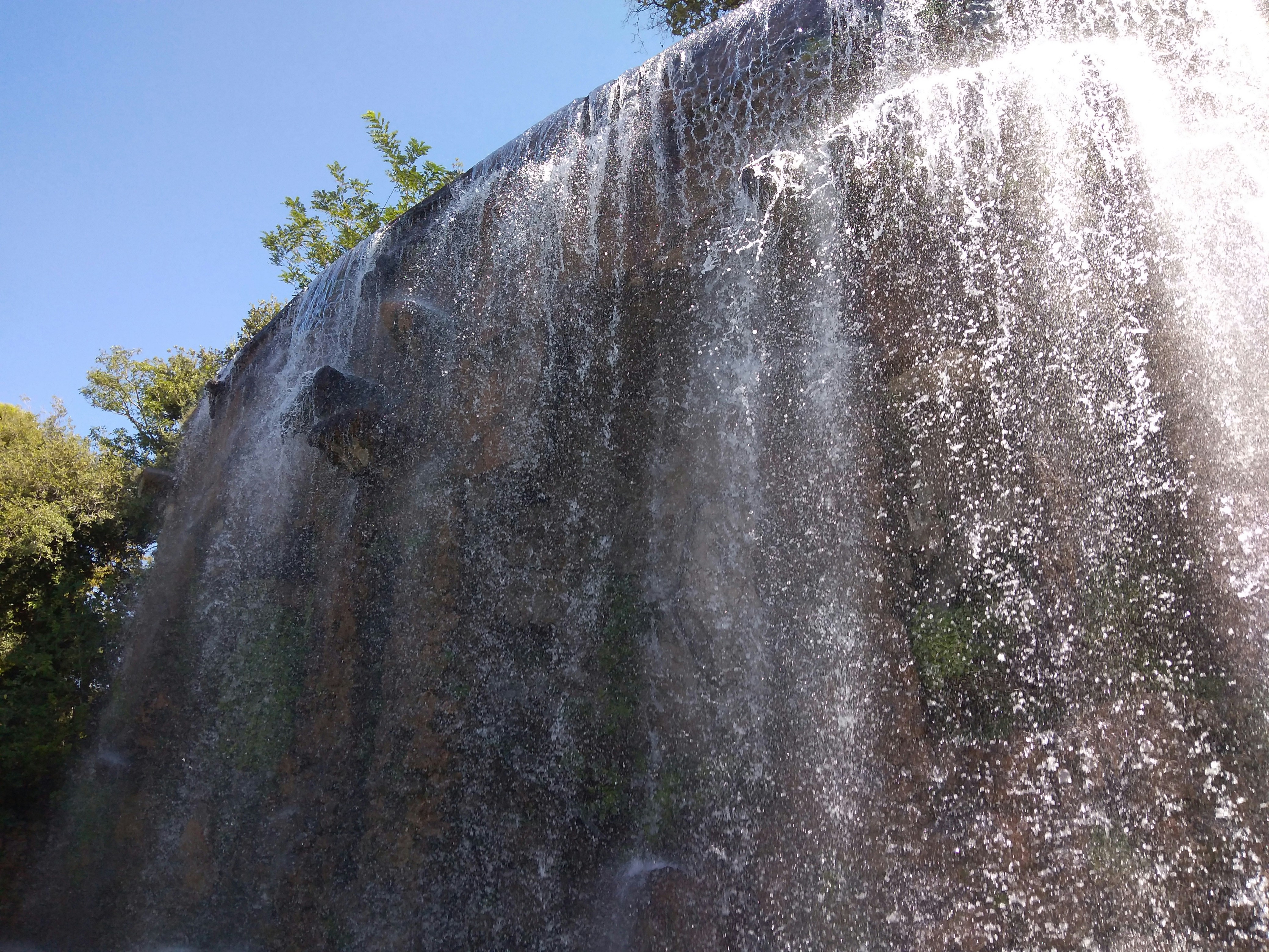 Majestic waterfall cascading down rocky cliffs, surrounded by lush greenery under a clear blue sky.