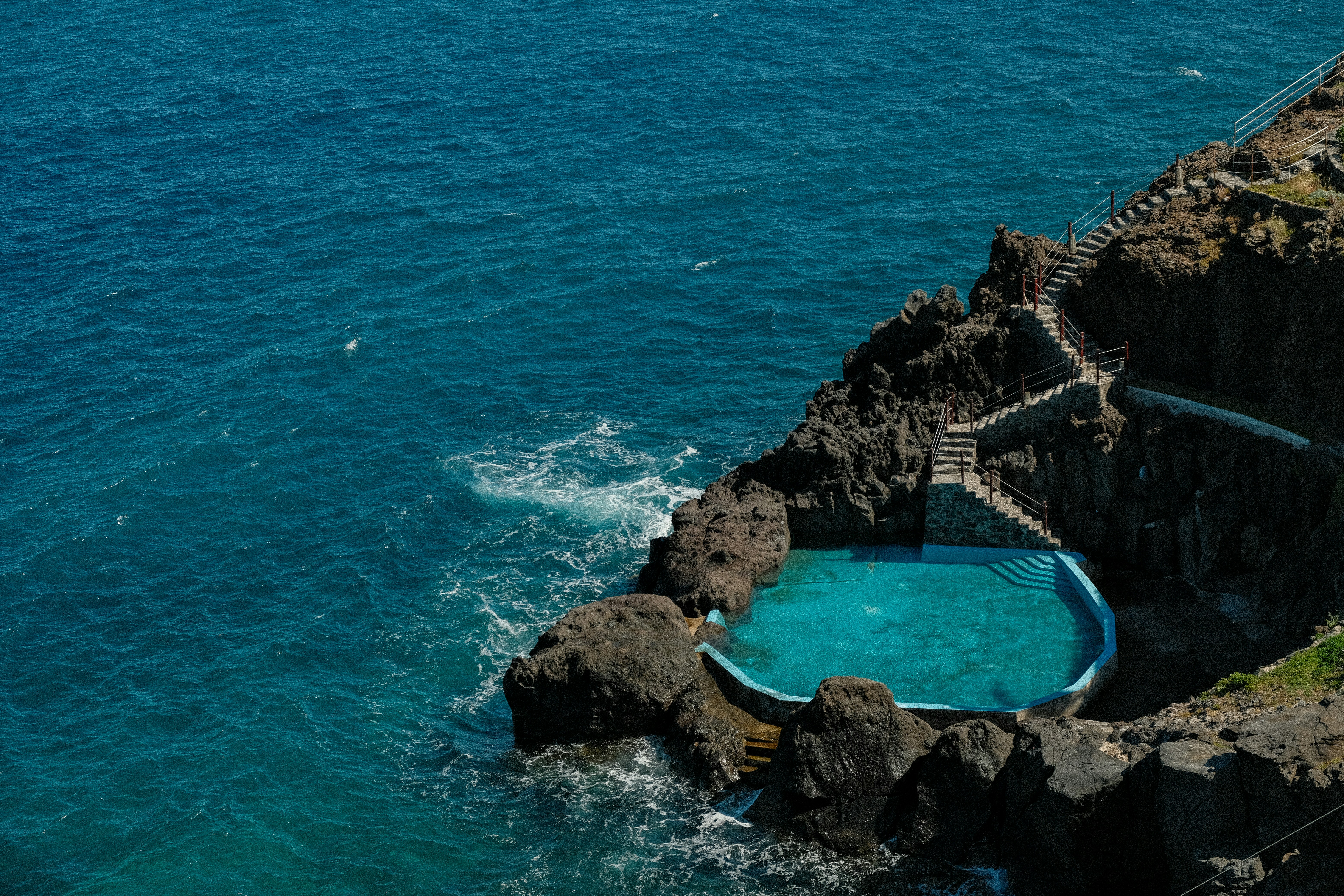 A tranquil swimming pool nestled among rugged rocks, with waves gently crashing against the shore. The vibrant blue water contrasts beautifully with the deep ocean backdrop.