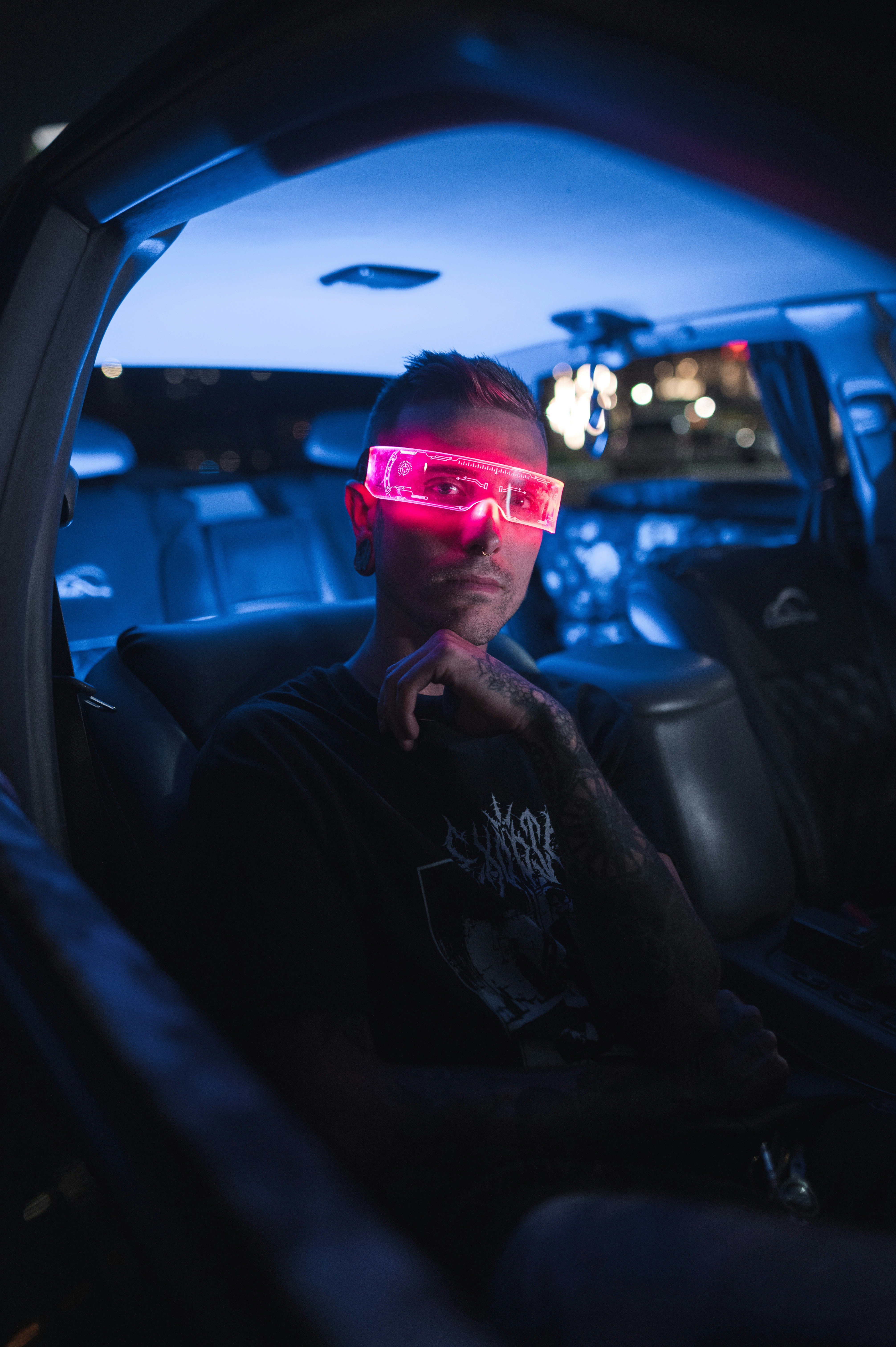 A person with vibrant pink glasses sits inside a car, illuminated by blue ambient light, embodying a modern urban aesthetic.