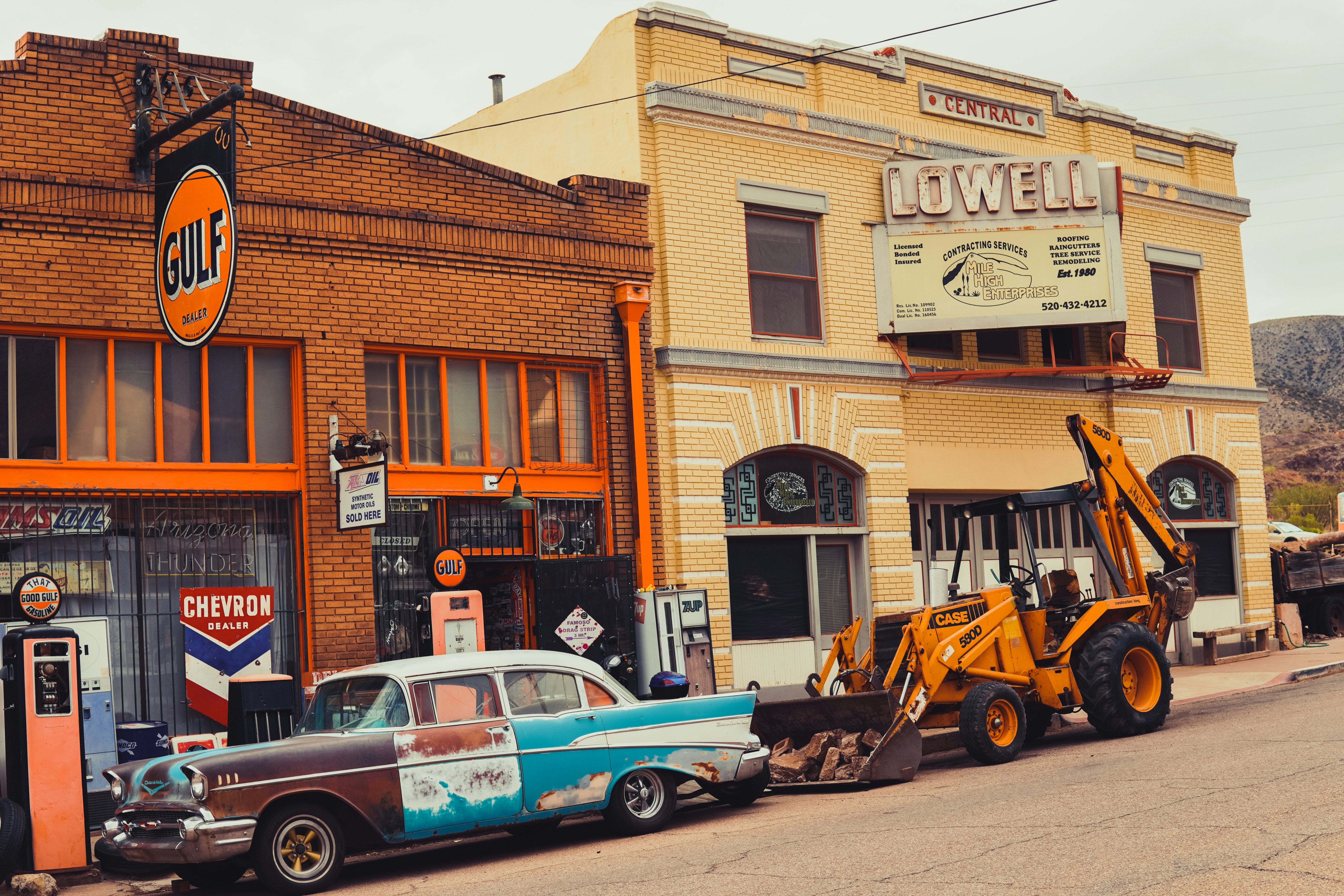 Vintage car and construction vehicle parked in front of historic buildings with retro signage.