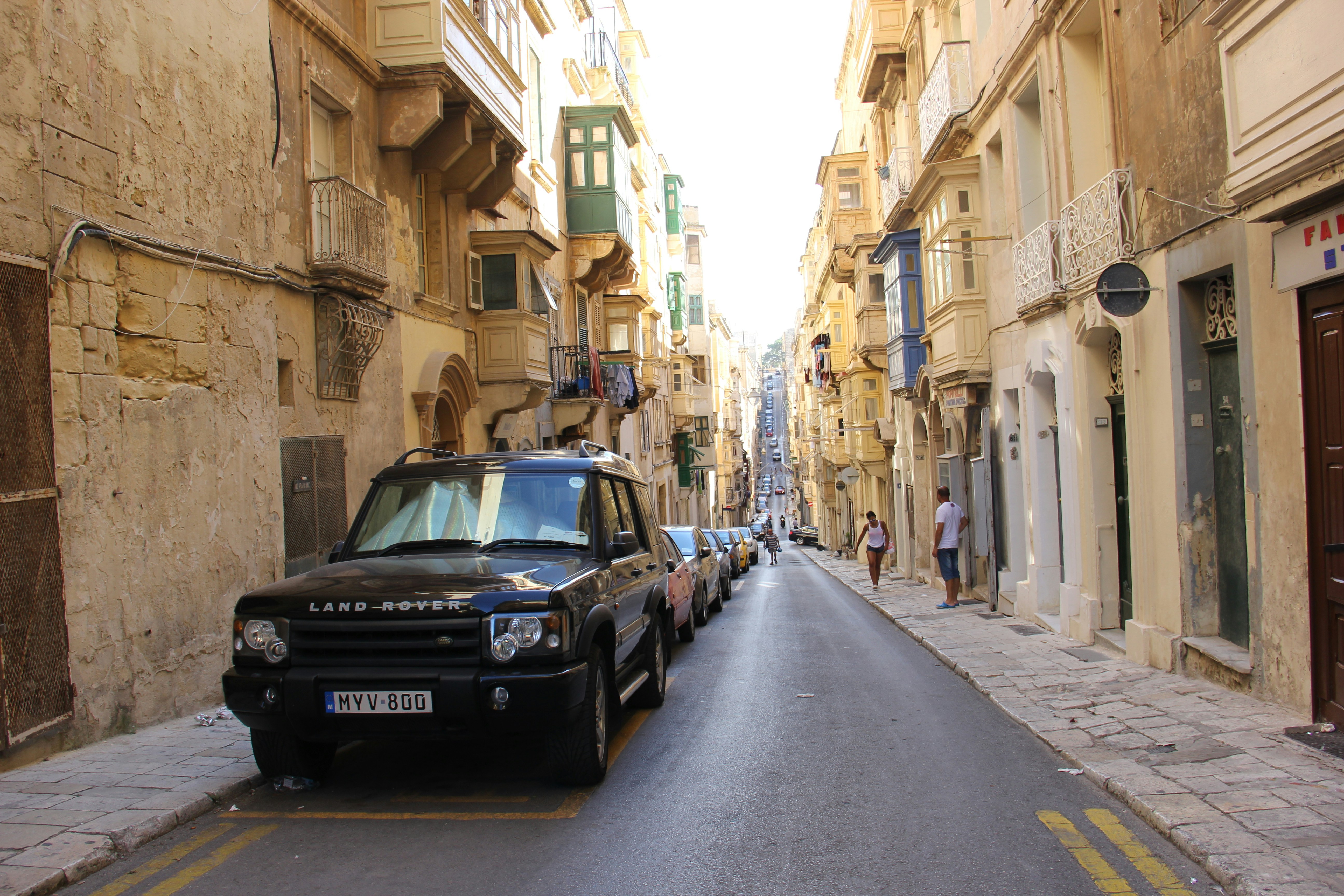 Narrow cobblestone street in Valletta, Malta, lined with parked cars and traditional architecture. A lone pedestrian strolls amidst the vibrant urban setting.