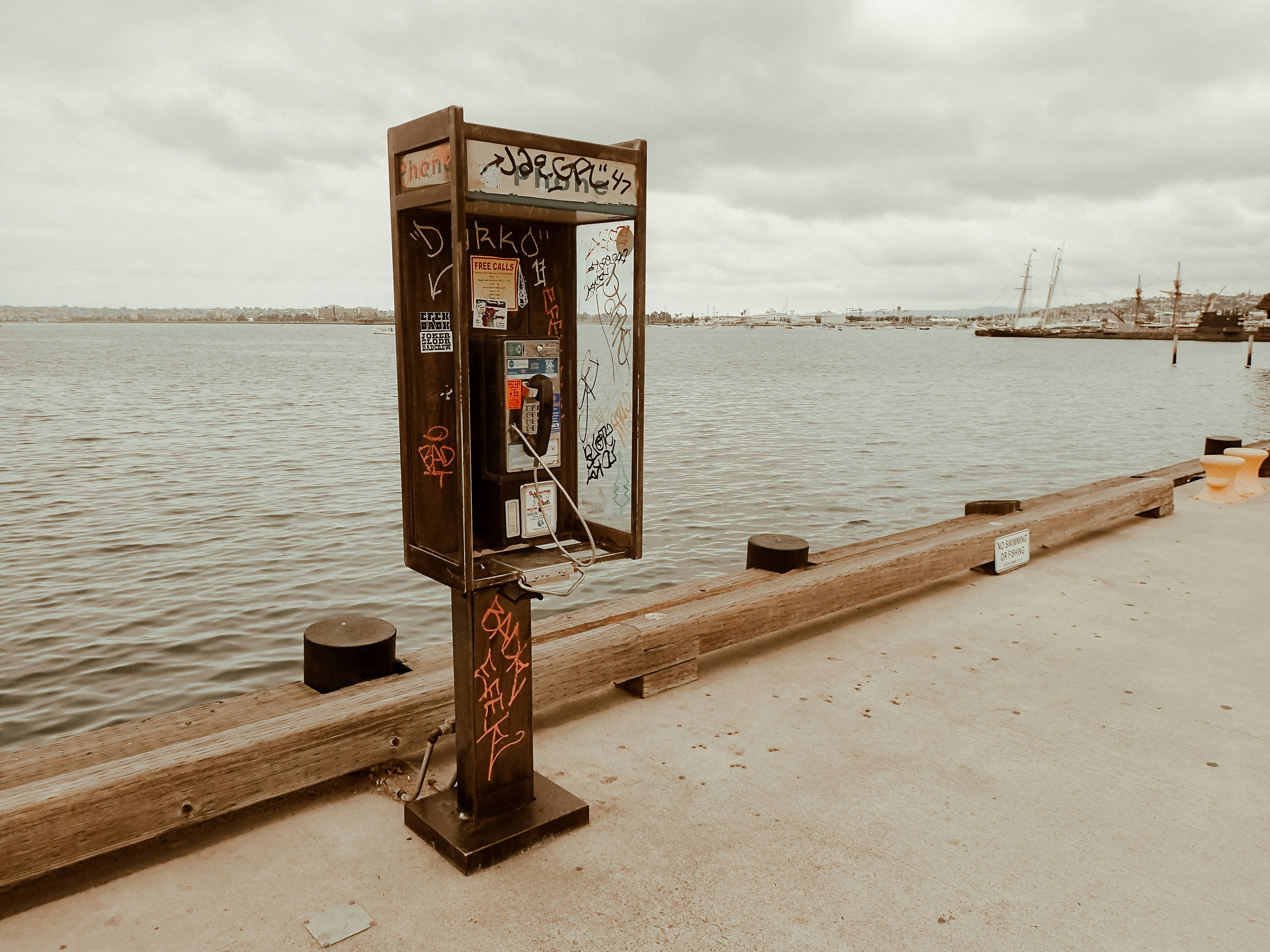 A weathered graffiti-covered payphone frame on a concrete promenade beside a calm harbor, with a distant shoreline.