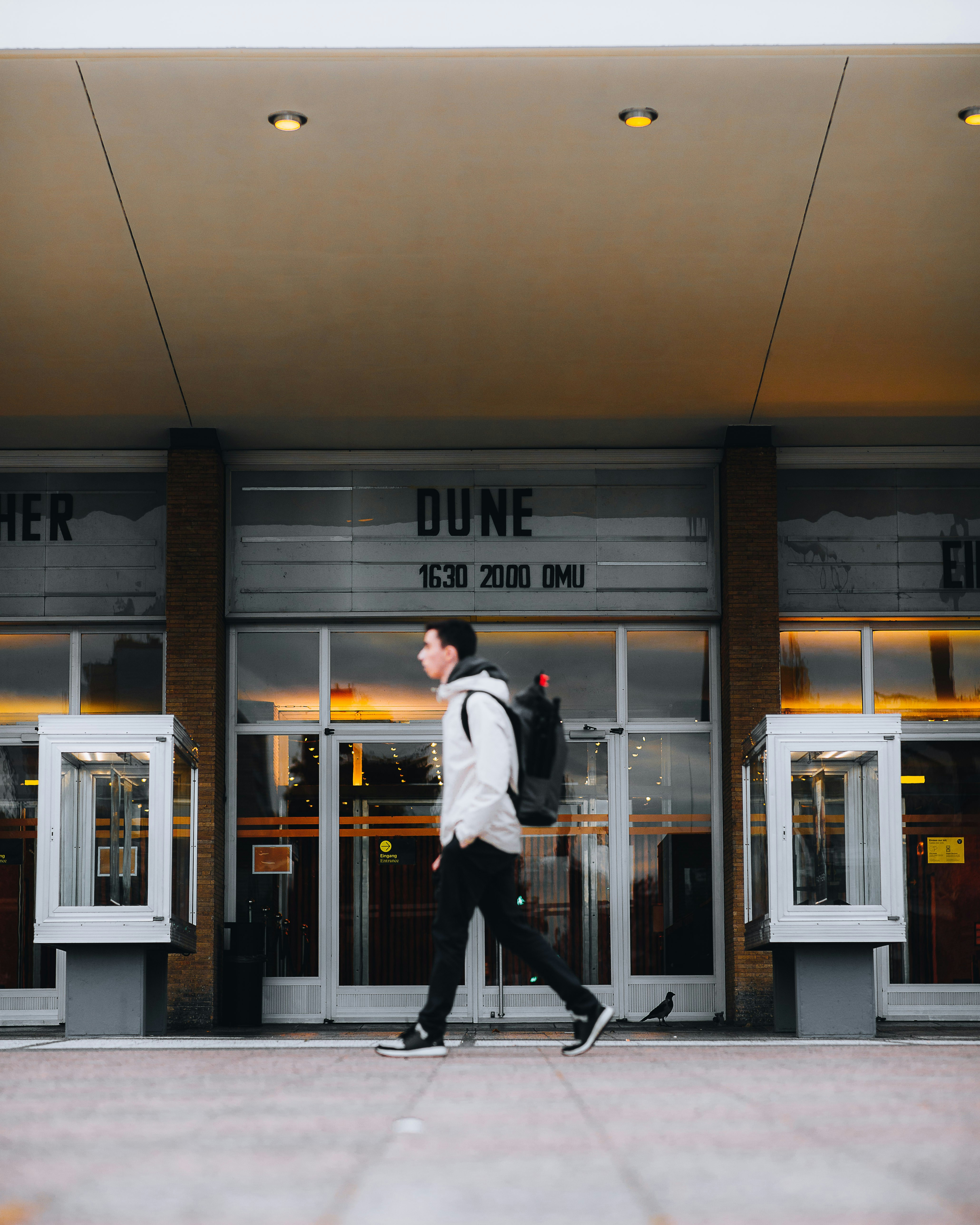 DUNE Under Neon: A solitary traveler strides past a glass-fronted storefront
