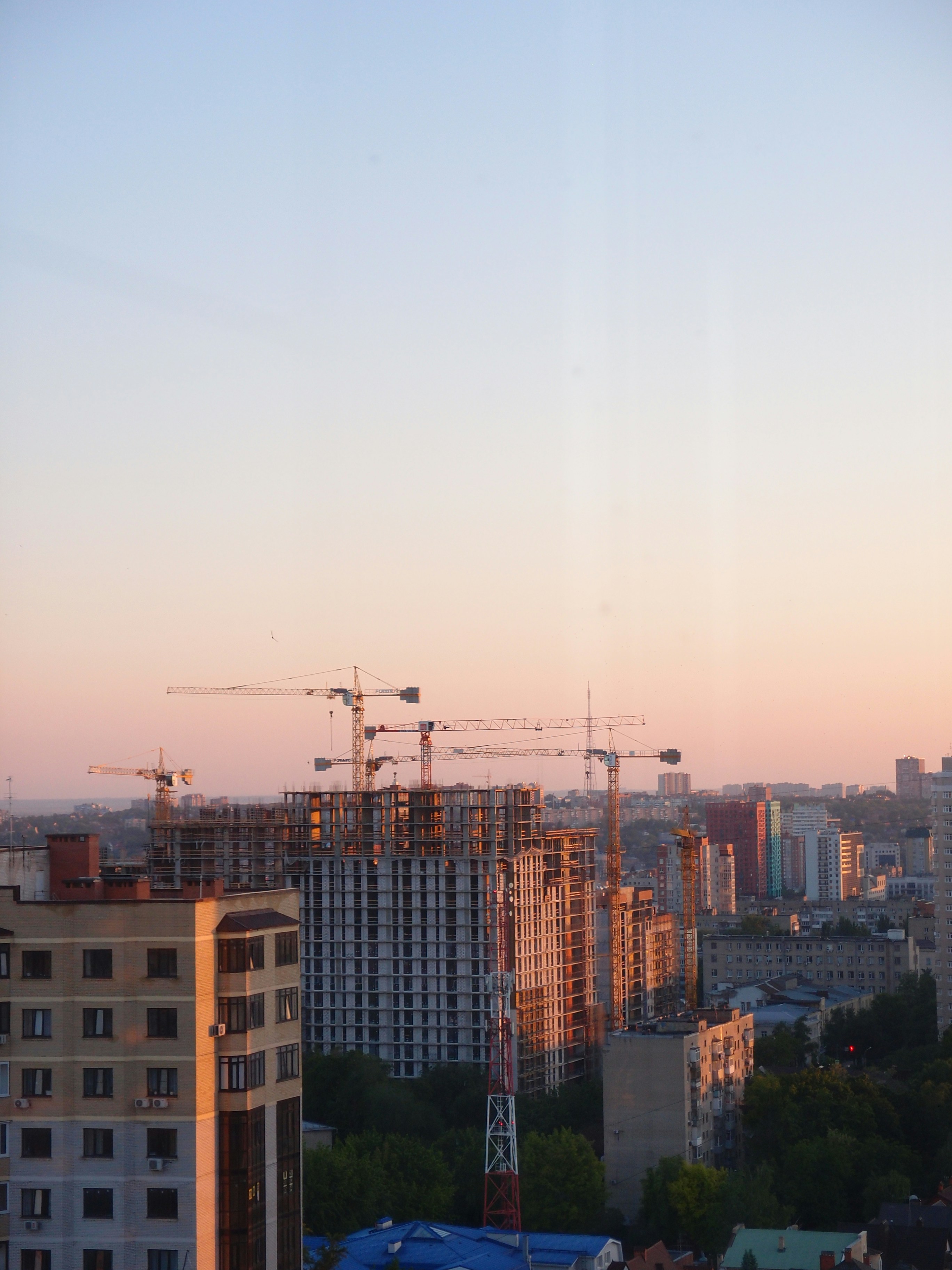 City skyline at dusk with several construction cranes rising above mid-rise buildings. Warm light brushes the façades, emphasizing vertical lines and urban growth.