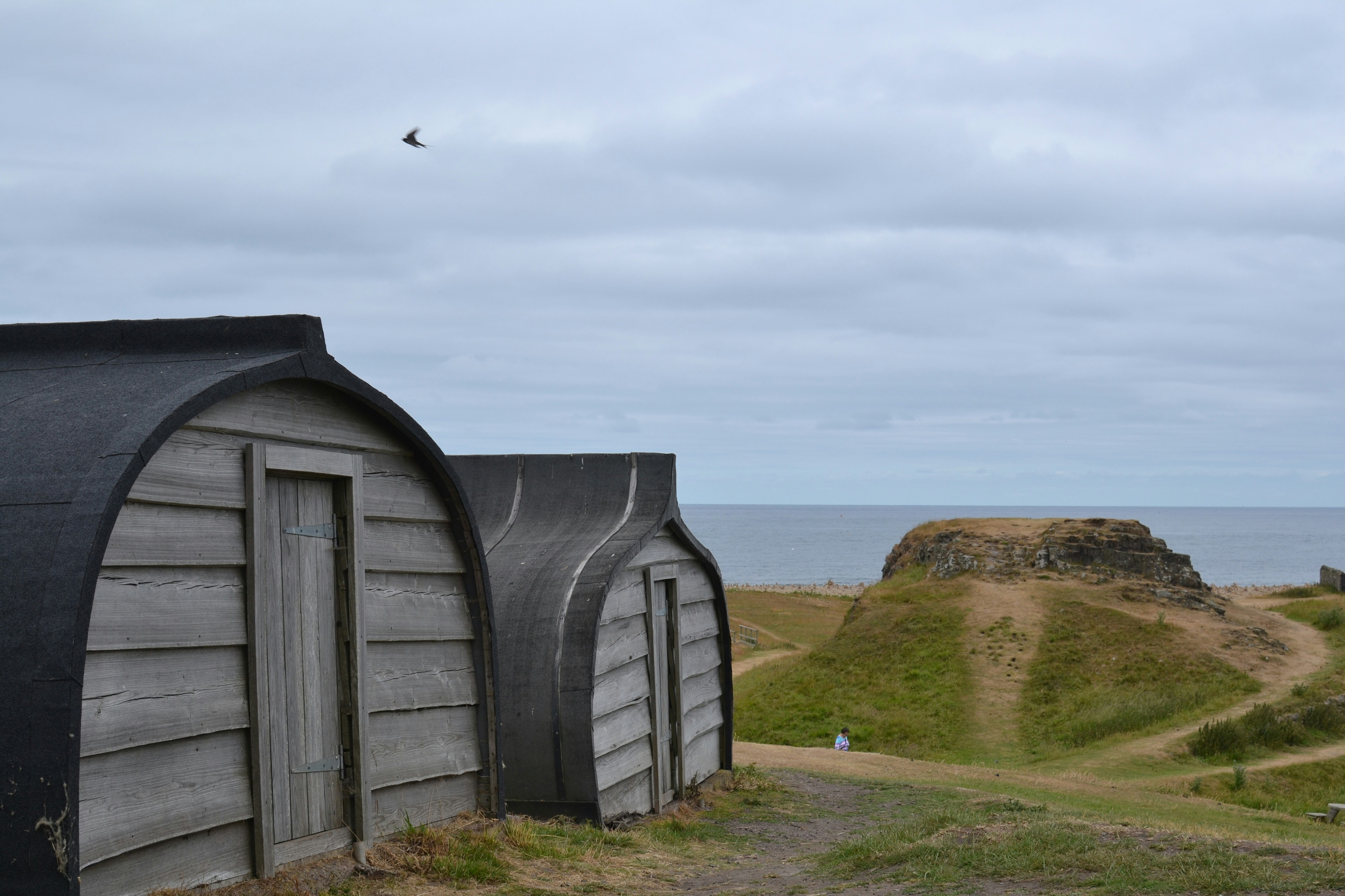 Two weathered structures stand sentinel near a coastal bluff, overlooking the calm sea under a cloudy sky.