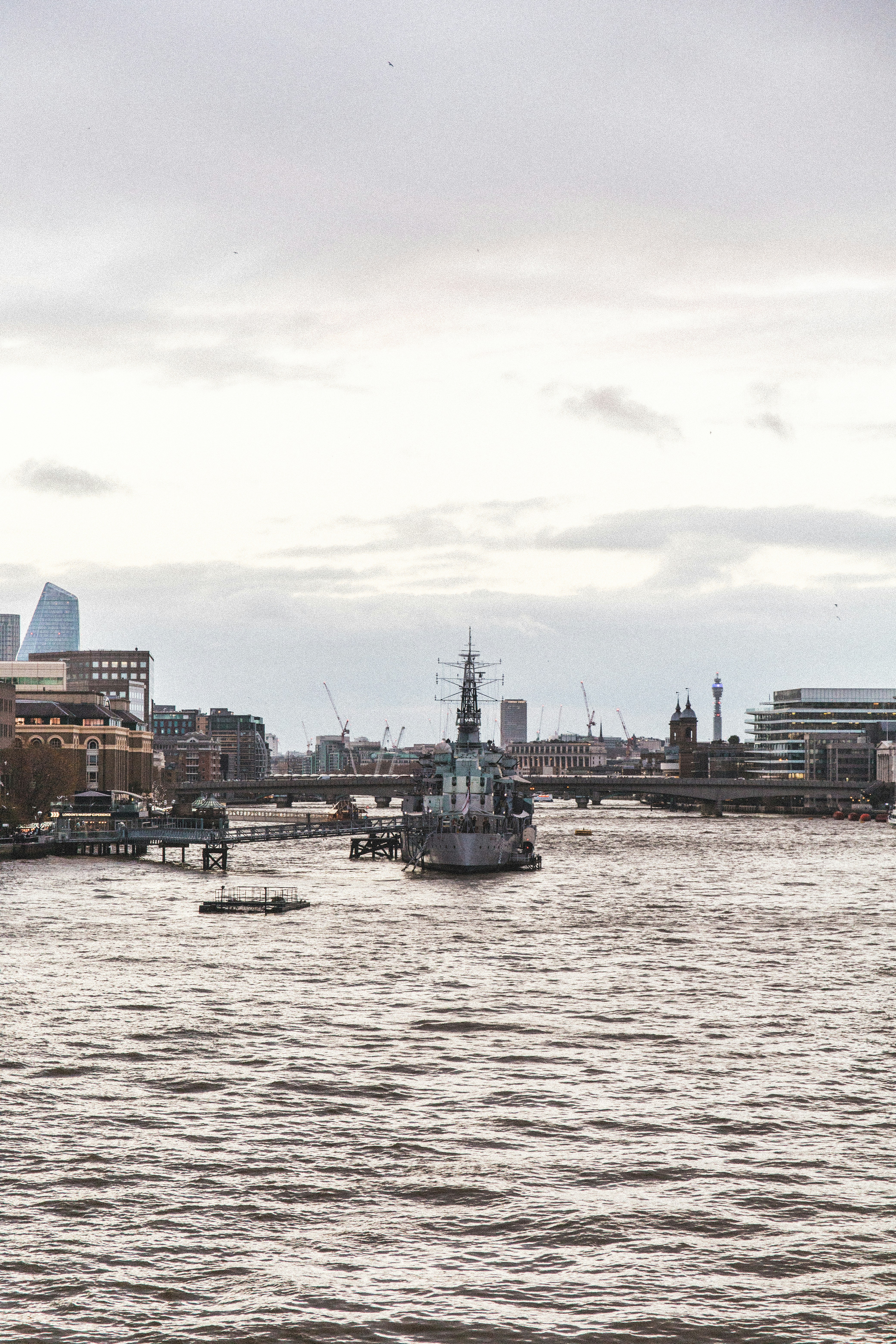 Historic ship anchored in the Thames River with modern cityscape in the background. Cloudy sky adds a dramatic ambiance.