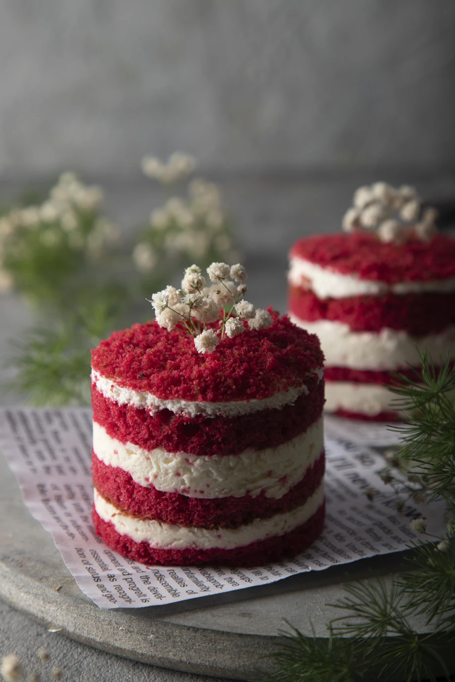 Two small red velvet layer cakes with white cream filling sit on printed paper, topped with tiny white flowers. One cake is in sharp focus in the foreground, with a second softly blurred behind it.