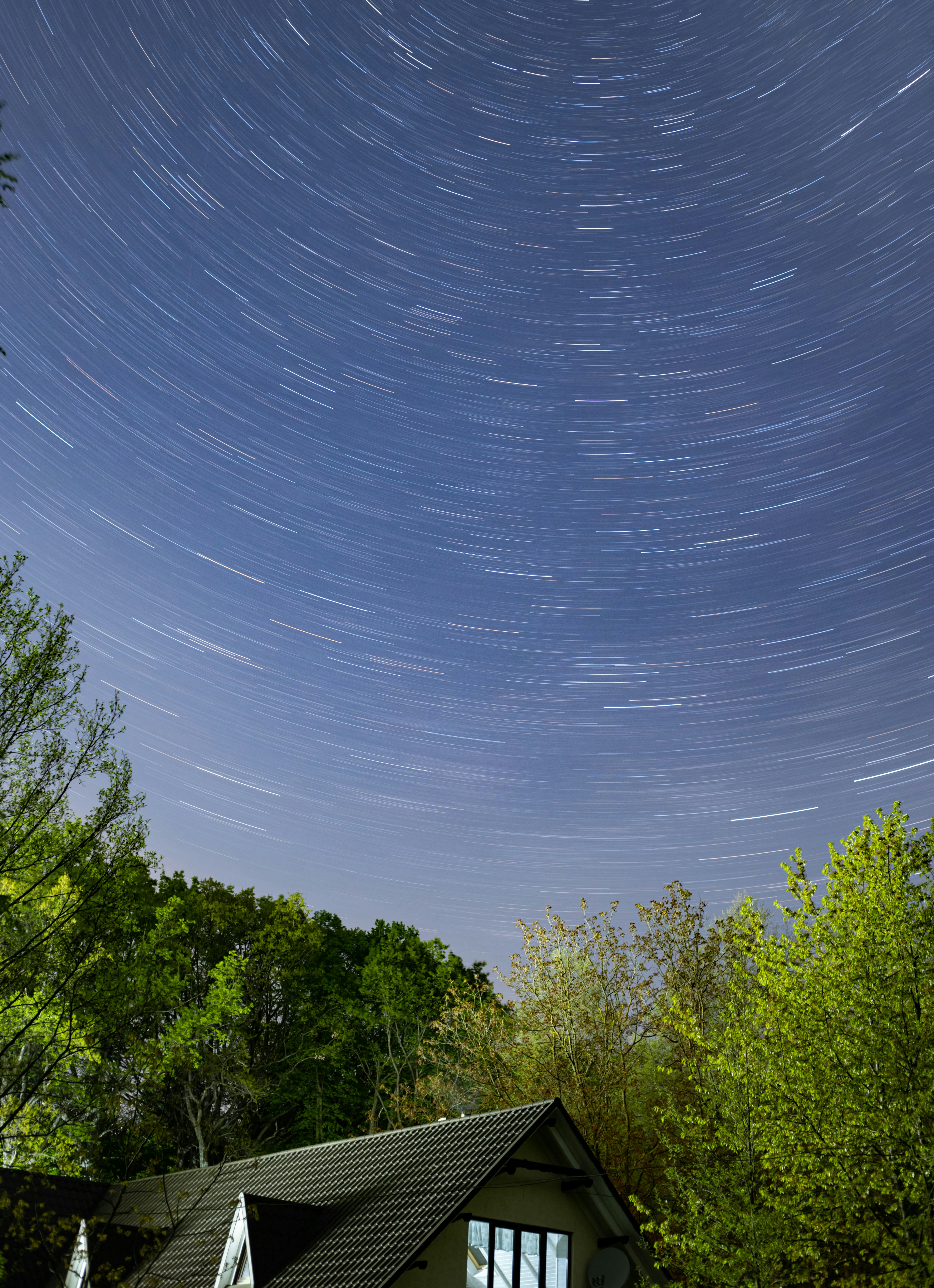 Star trails spiral above a tranquil house, framed by lush trees in a nighttime setting.