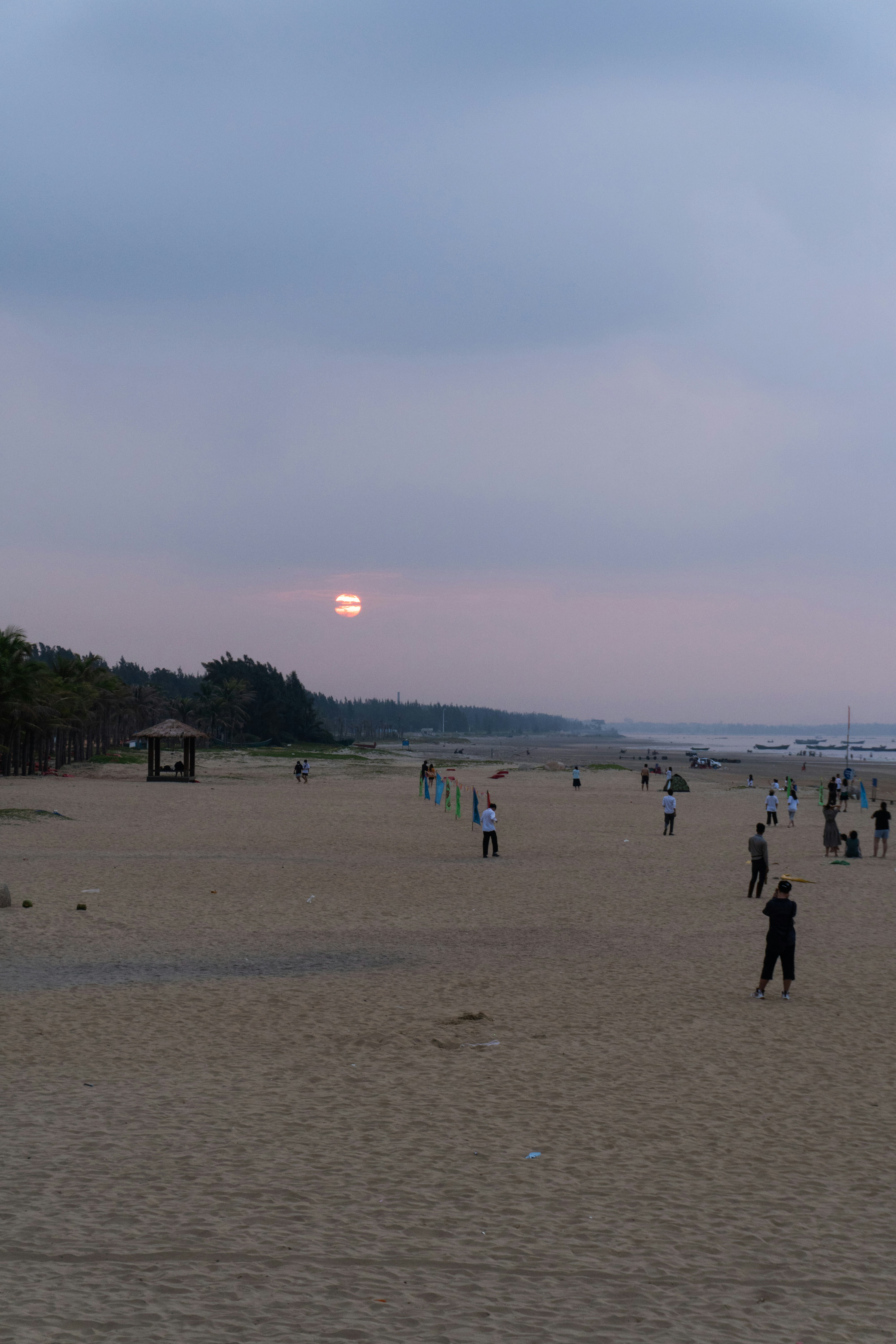 Beachgoers enjoy a tranquil evening as the sun sets behind a veil of clouds, casting a warm glow over the sandy expanse.