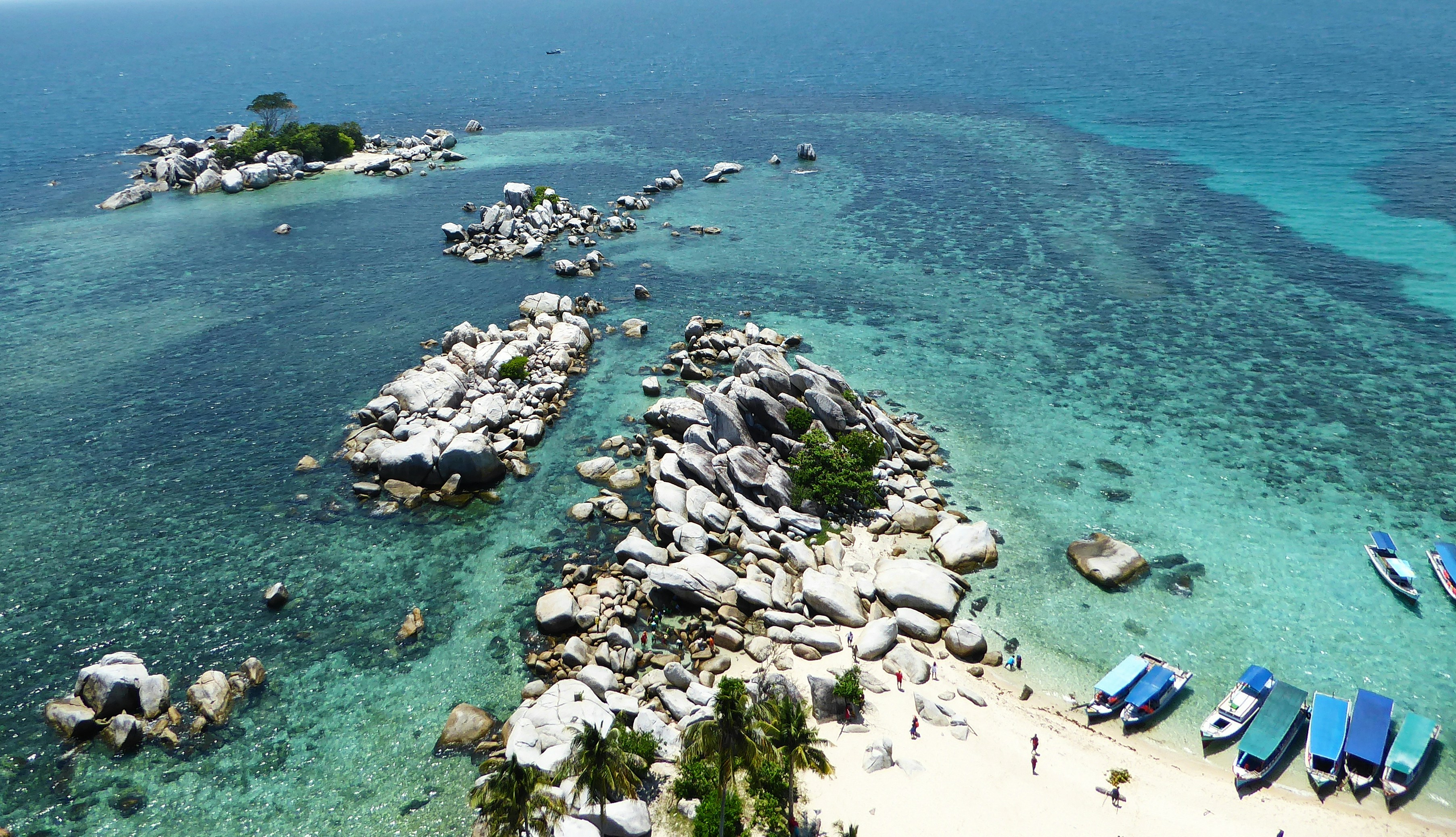 Aerial view of a tropical beach with rocky outcrops and vibrant turquoise waters, dotted with boats and lush greenery.