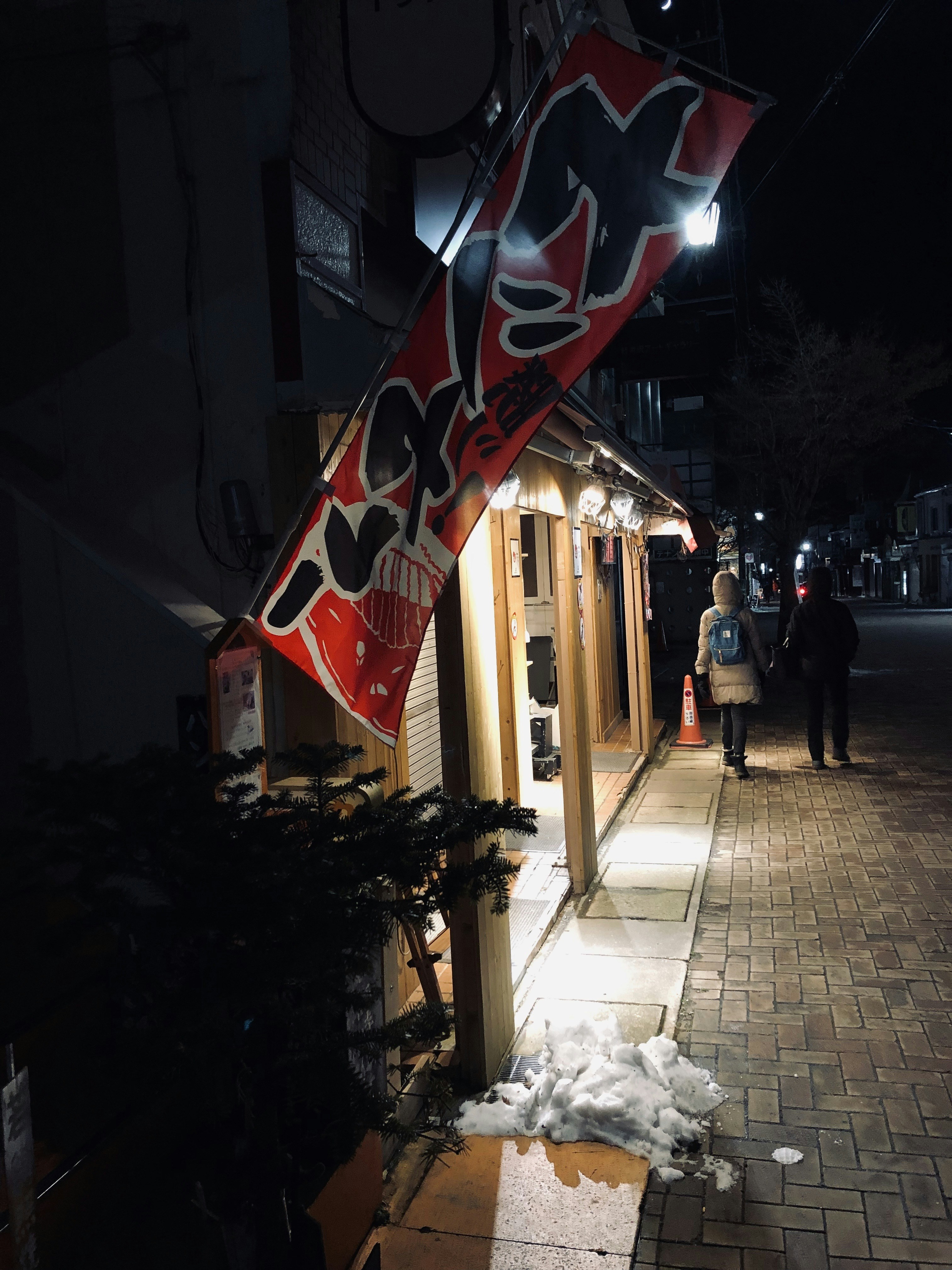 A vibrant red banner flutters outside a cozy eatery, illuminated by warm lights, as pedestrians stroll down a quiet, snowy street at night.