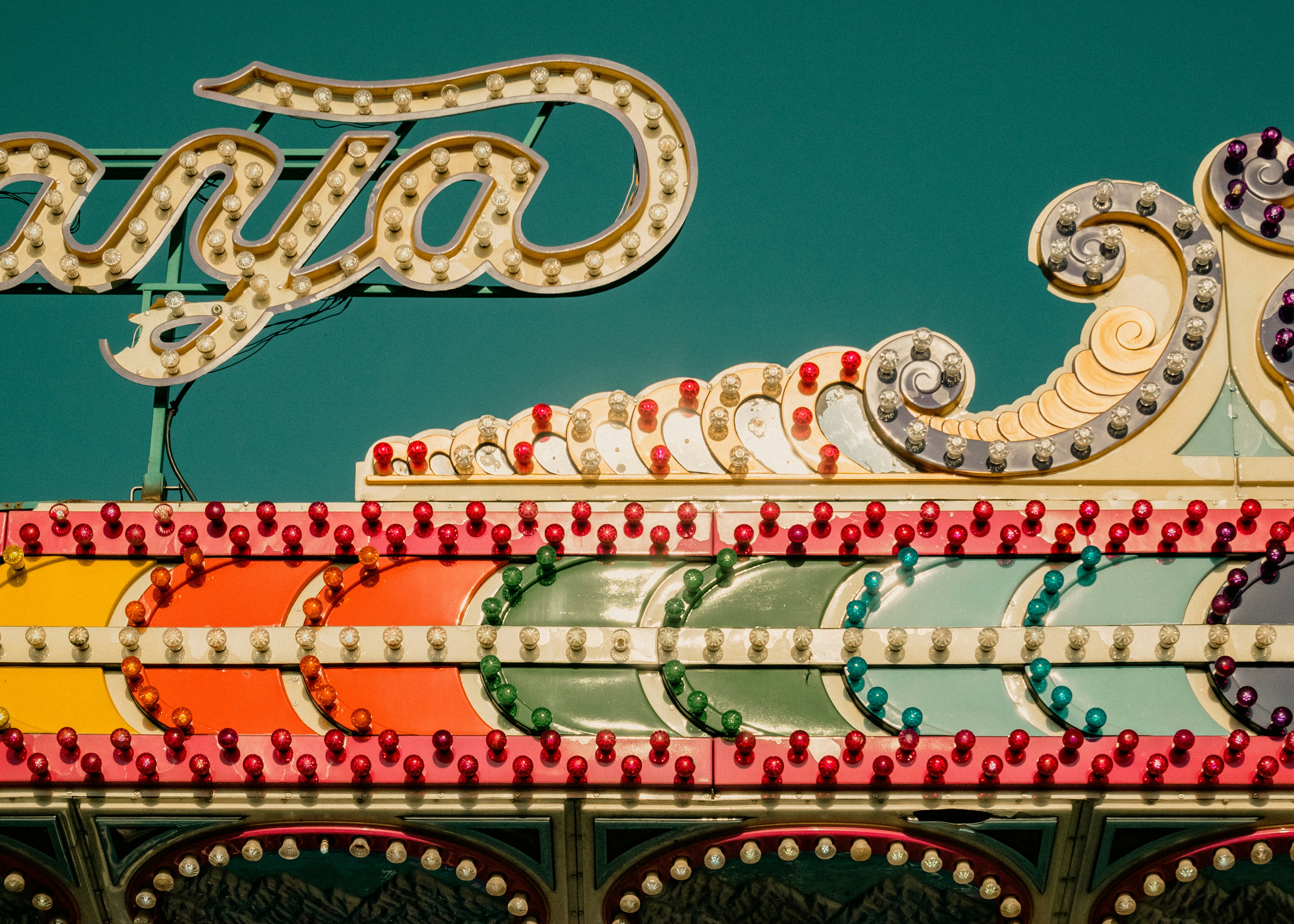 Close-up of a vintage carnival marquee adorned with multicolored bulbs and neon lettering against a teal sky. This photograph captures the decorative signage and festive glow.