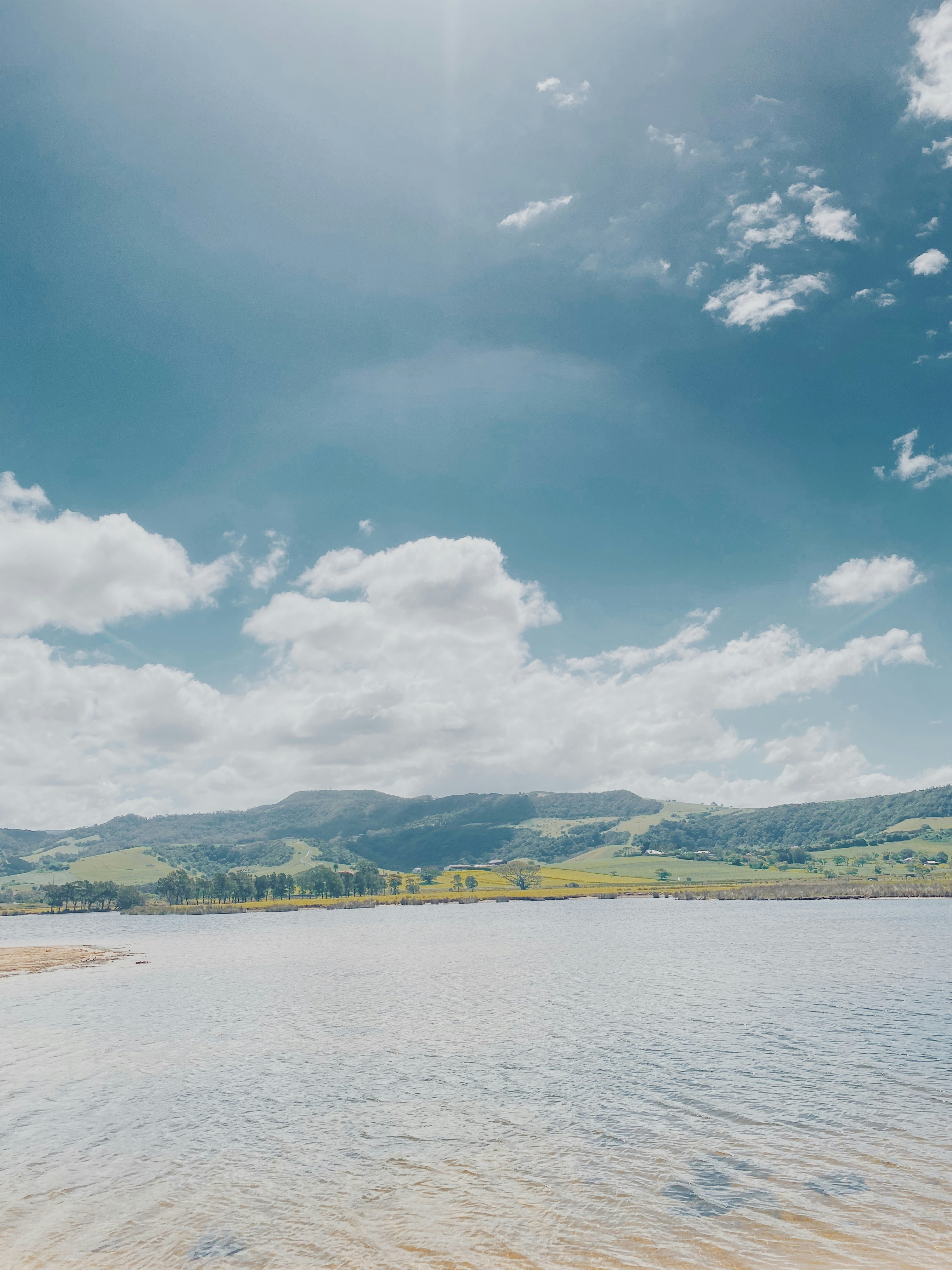 Expansive view of a serene lake bordered by lush hills and a vibrant sky dotted with clouds.