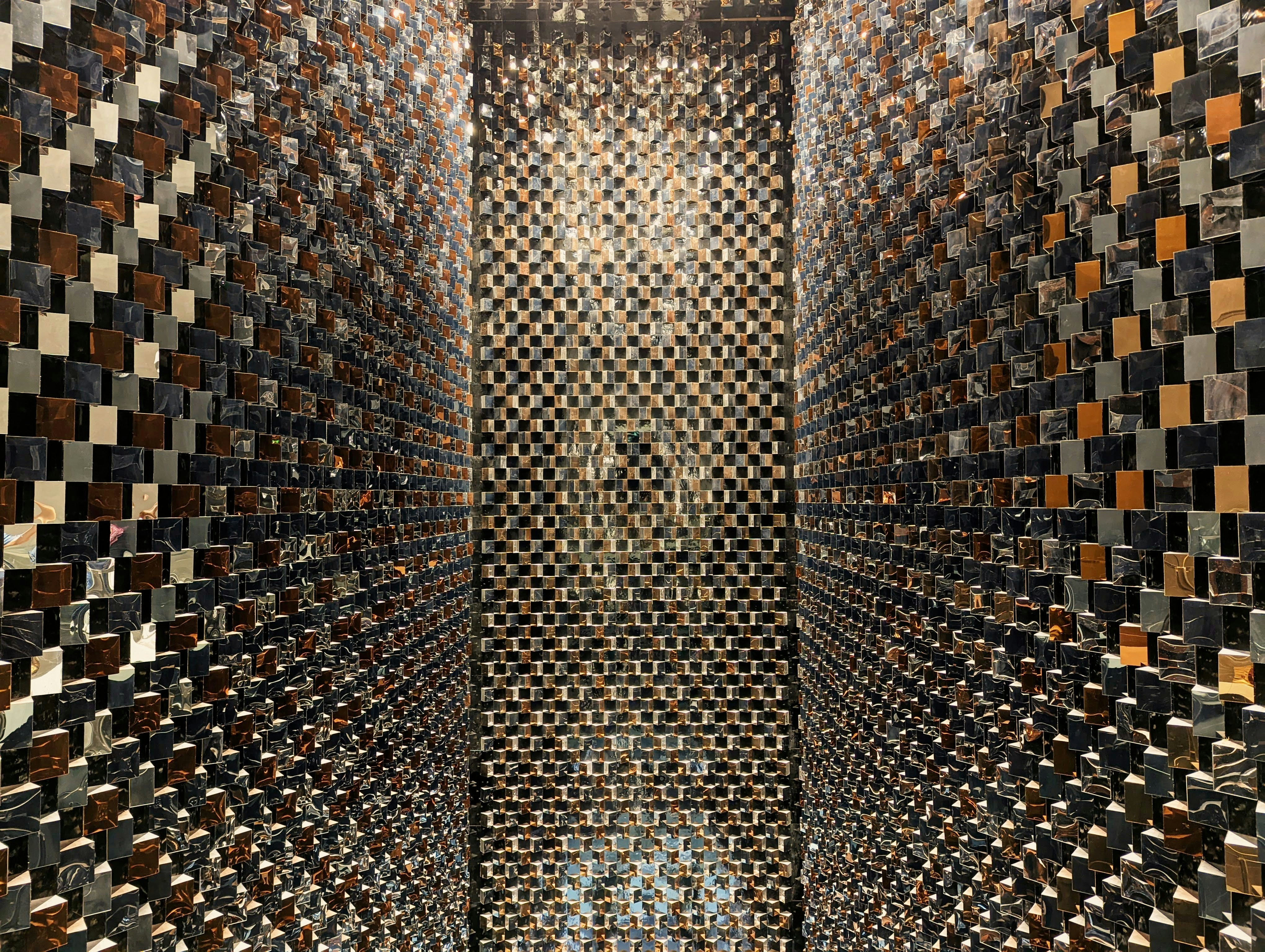 Narrow corridor lined with tiny square tiles in metallic tones, converging toward a glowing end.