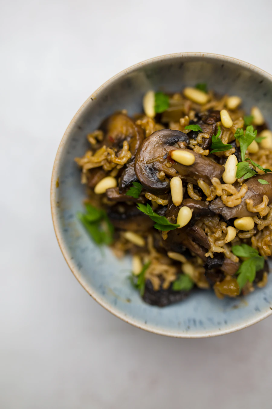 A ceramic bowl filled with sautéed mushrooms, grains, pine nuts, and fresh parsley on a white background.